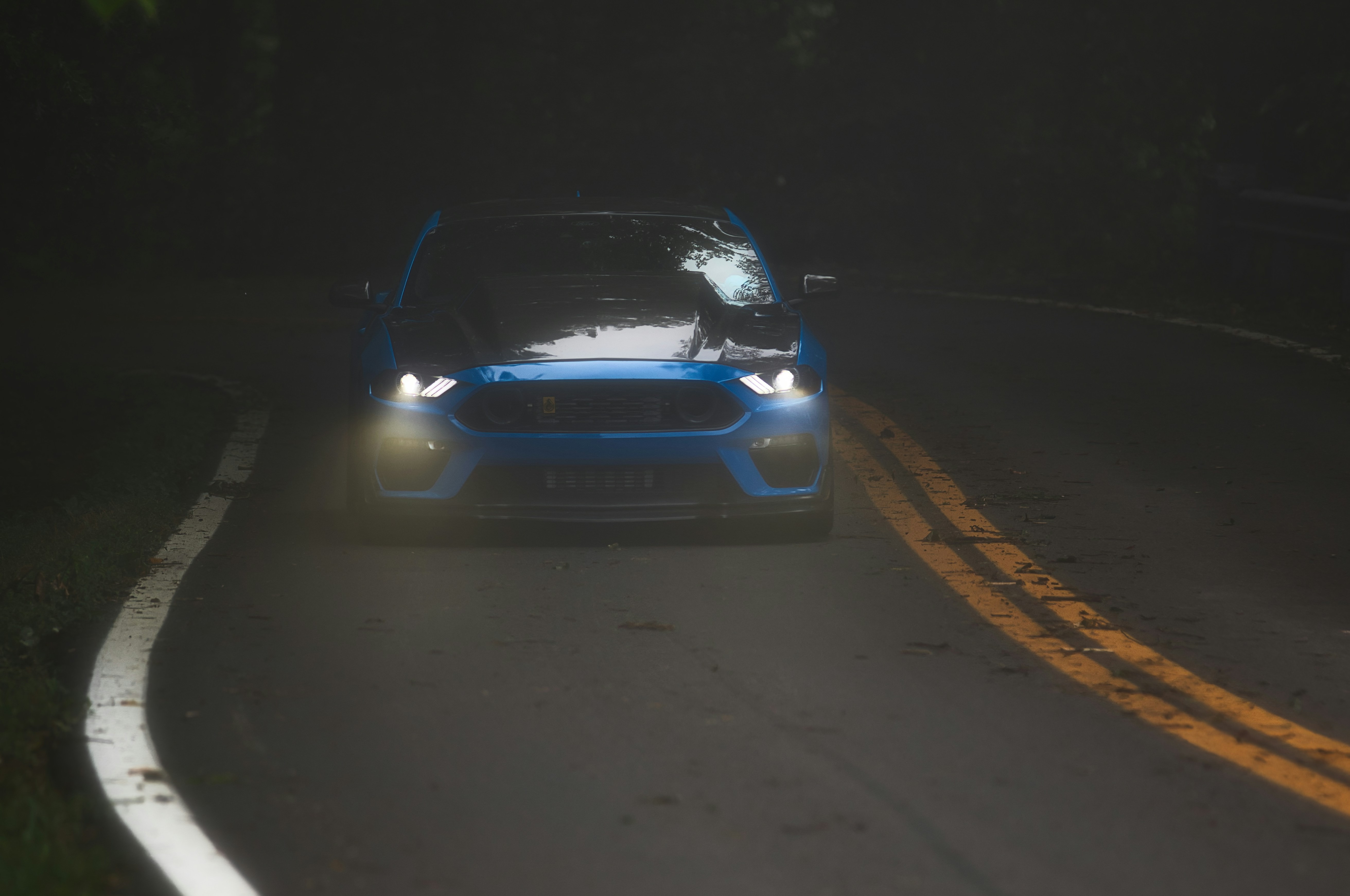 Blue rally-style car drives toward the camera on a dark, curved road; headlights cut through the low visibility, with white roadside markings and double yellow center lines visible.