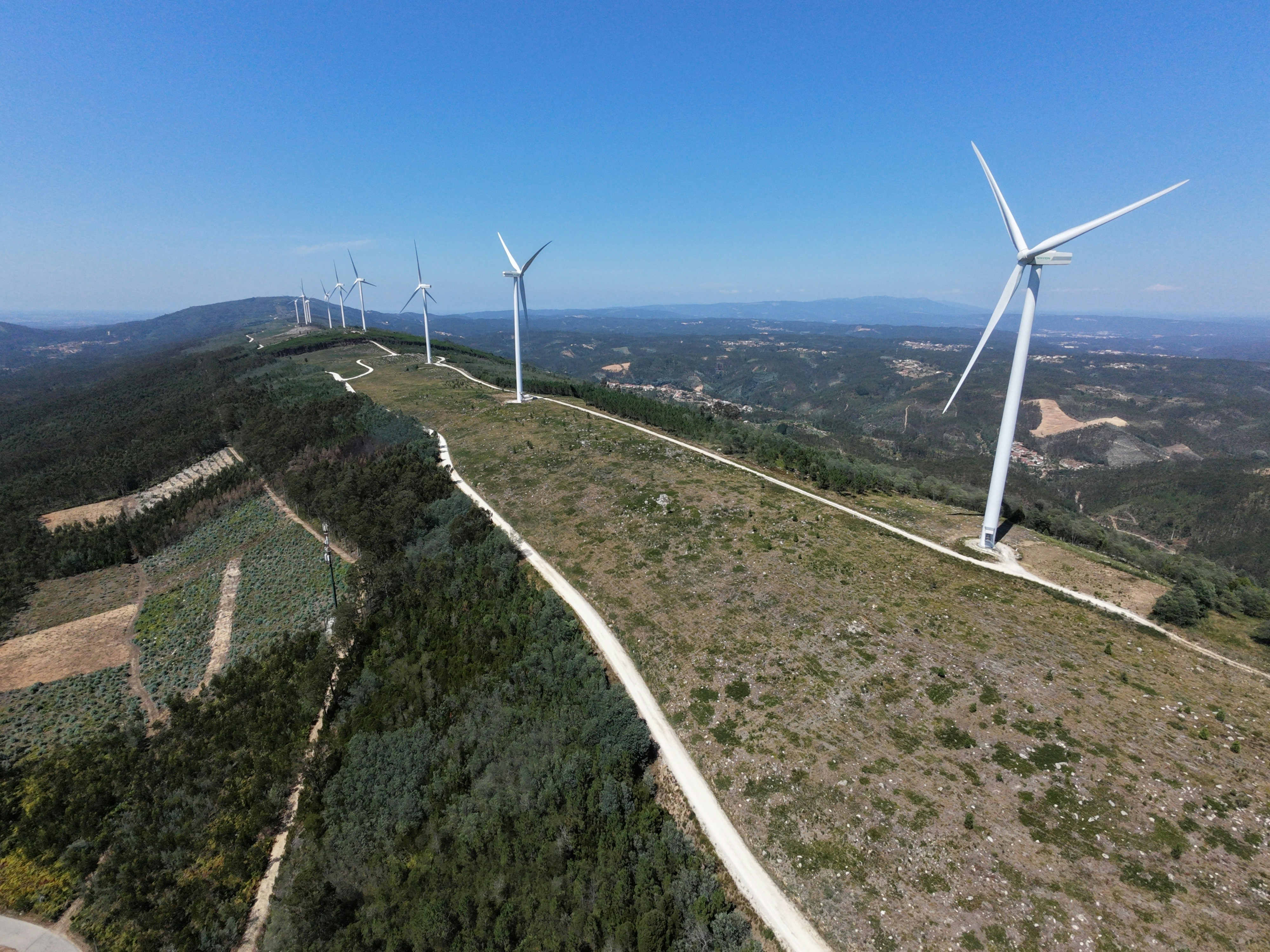 Aerial view of wind turbines along a mountain ridge with forests, dirt roads, and distant hills.