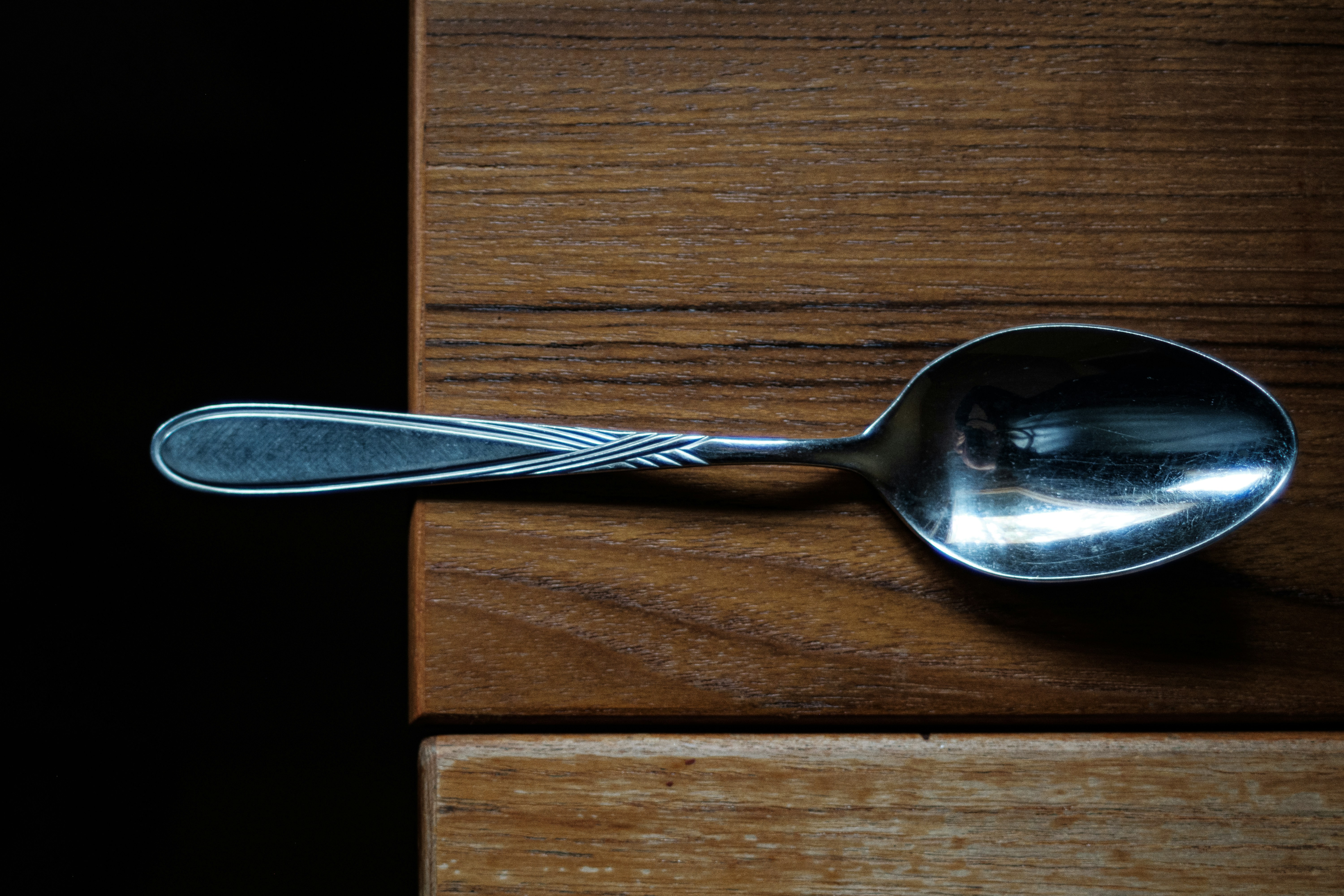 Shiny metal spoon lies along the seam of a wooden surface, capturing reflections on its bowl; the grain and tone of the wood provide a warm contrast to the sleek metallic shape.