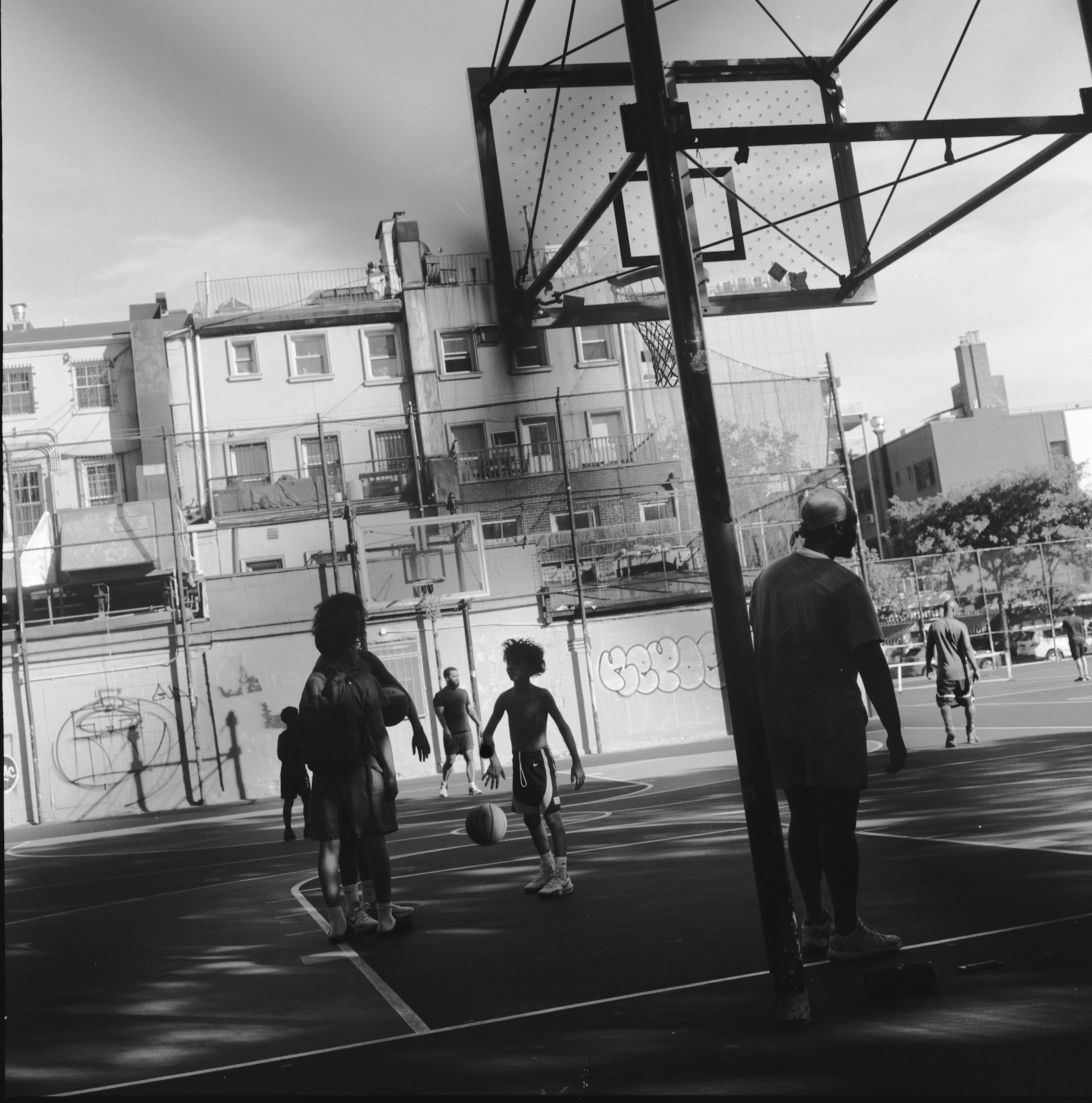 A grayscale street basketball court scene with children playing under a hoop while an adult watches; graffiti and urban buildings form the backdrop. This is a candid street photography frame.