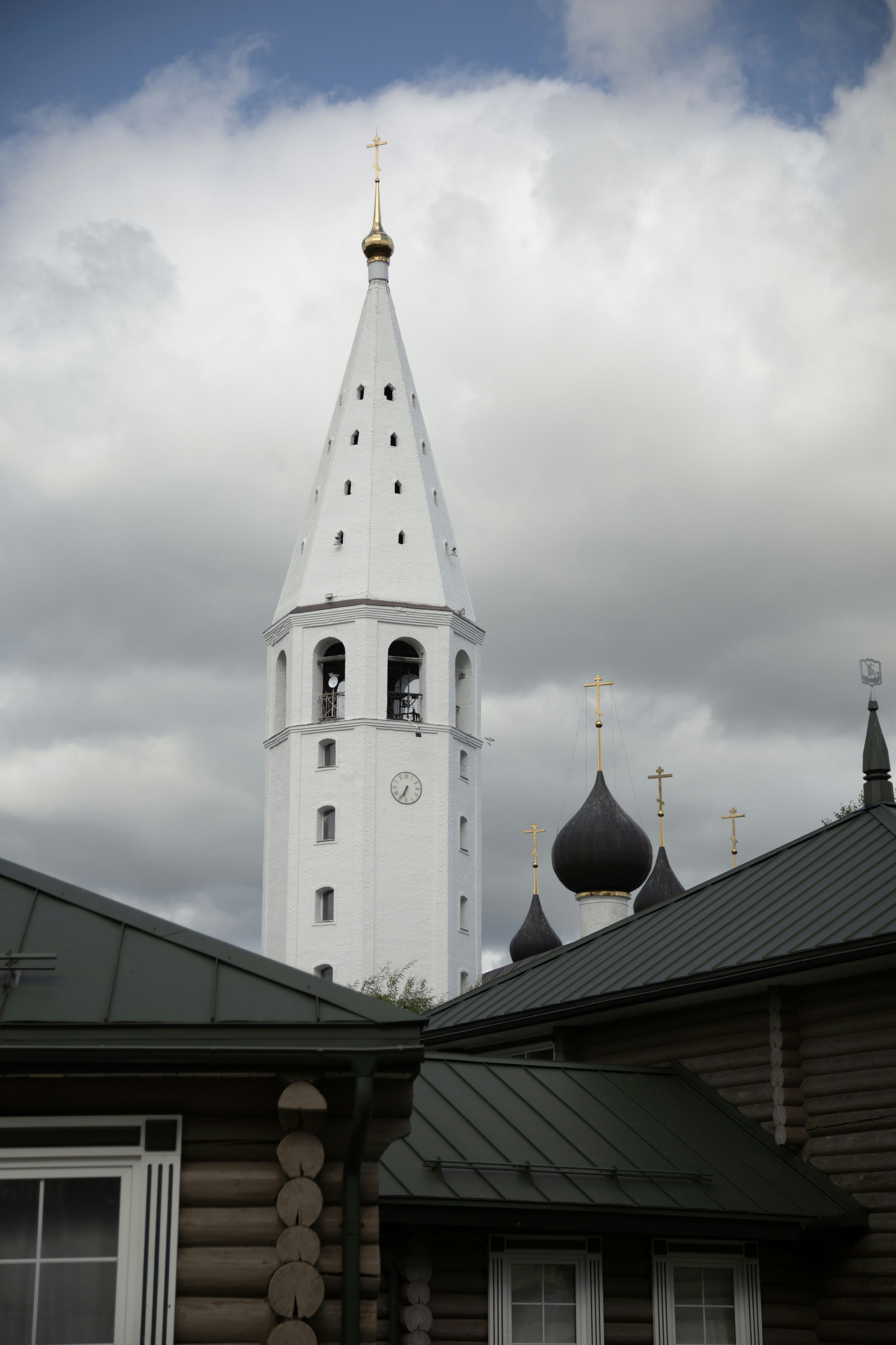 White Bell Tower Rising Above Green Rooftops Beneath A Cloudy Sky