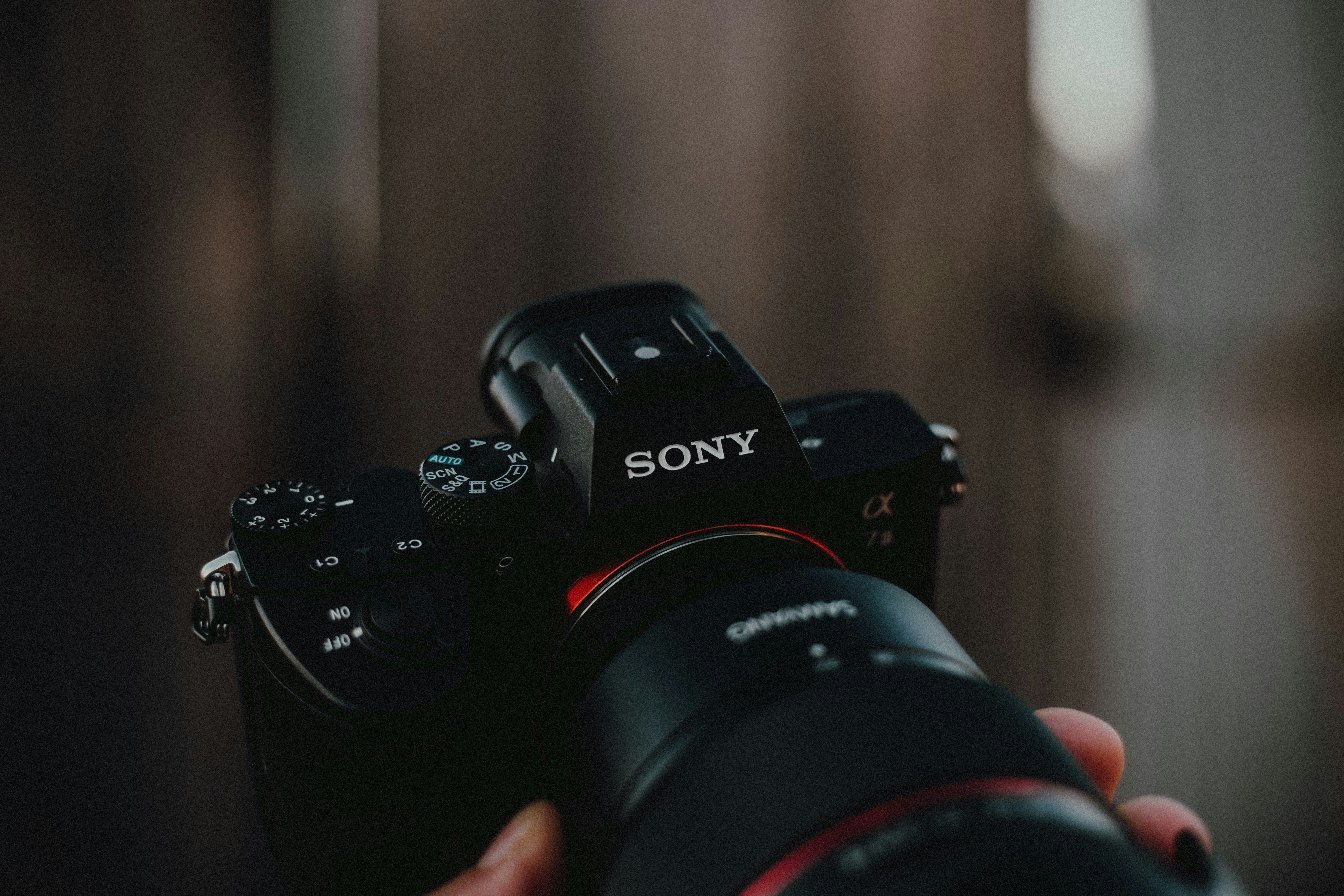 Close-up of a Sony Alpha camera with a red-ring lens, held in a hand against a dark, blurred background.