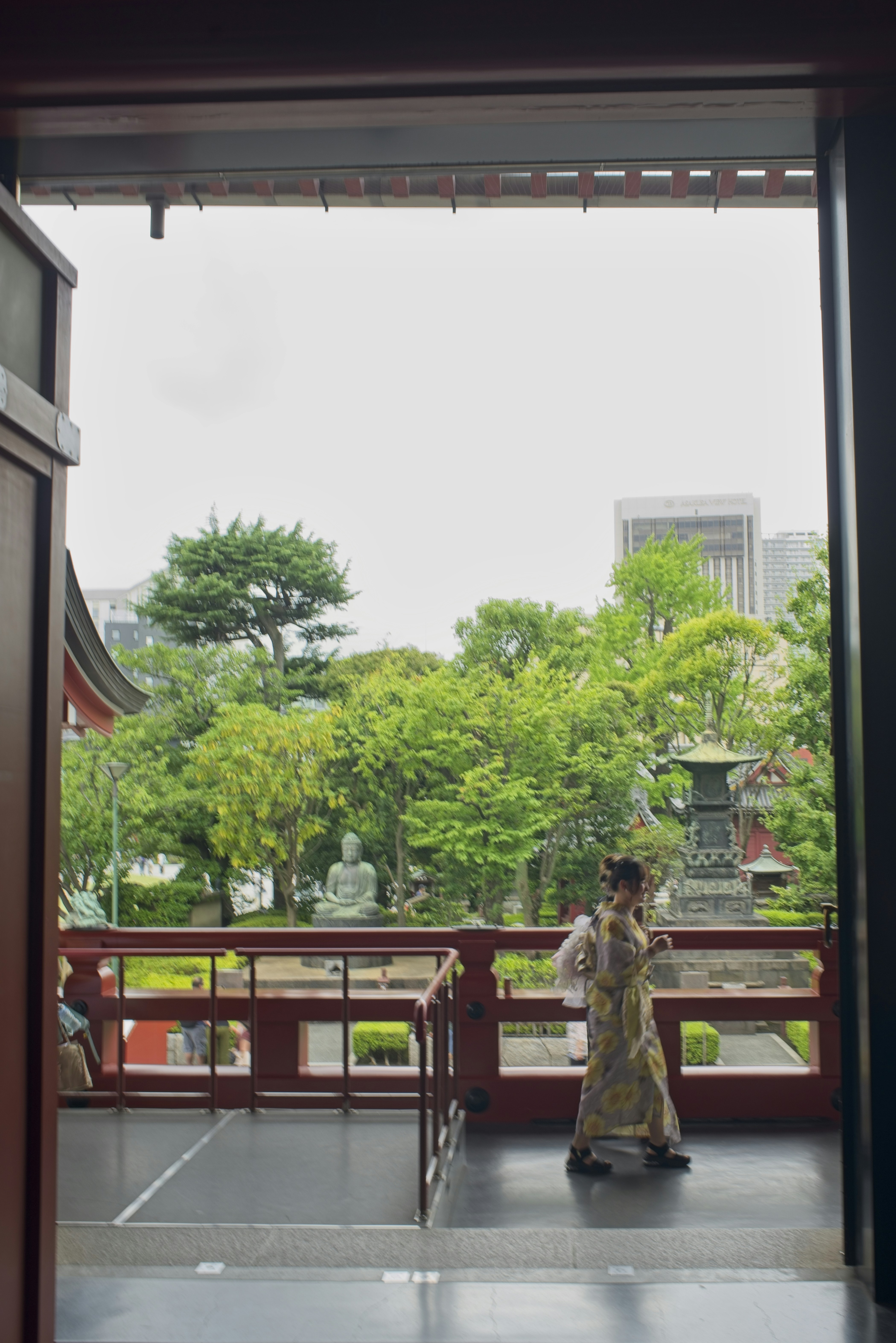 A woman in a floral kimono walks along Senso-ji's red temple balcony, framed by an open doorway that reveals a serene garden below. A seated bronze Buddha statue and an ornate lantern stand among lush green trees, with modern high-rises peeking through the foliage in the distance. This layered composition beautifully juxtaposes traditional temple architecture and dress against Tokyo's contemporary skyline, capturing a quiet moment within Asakusa's historic temple grounds on a warm July afternoon.