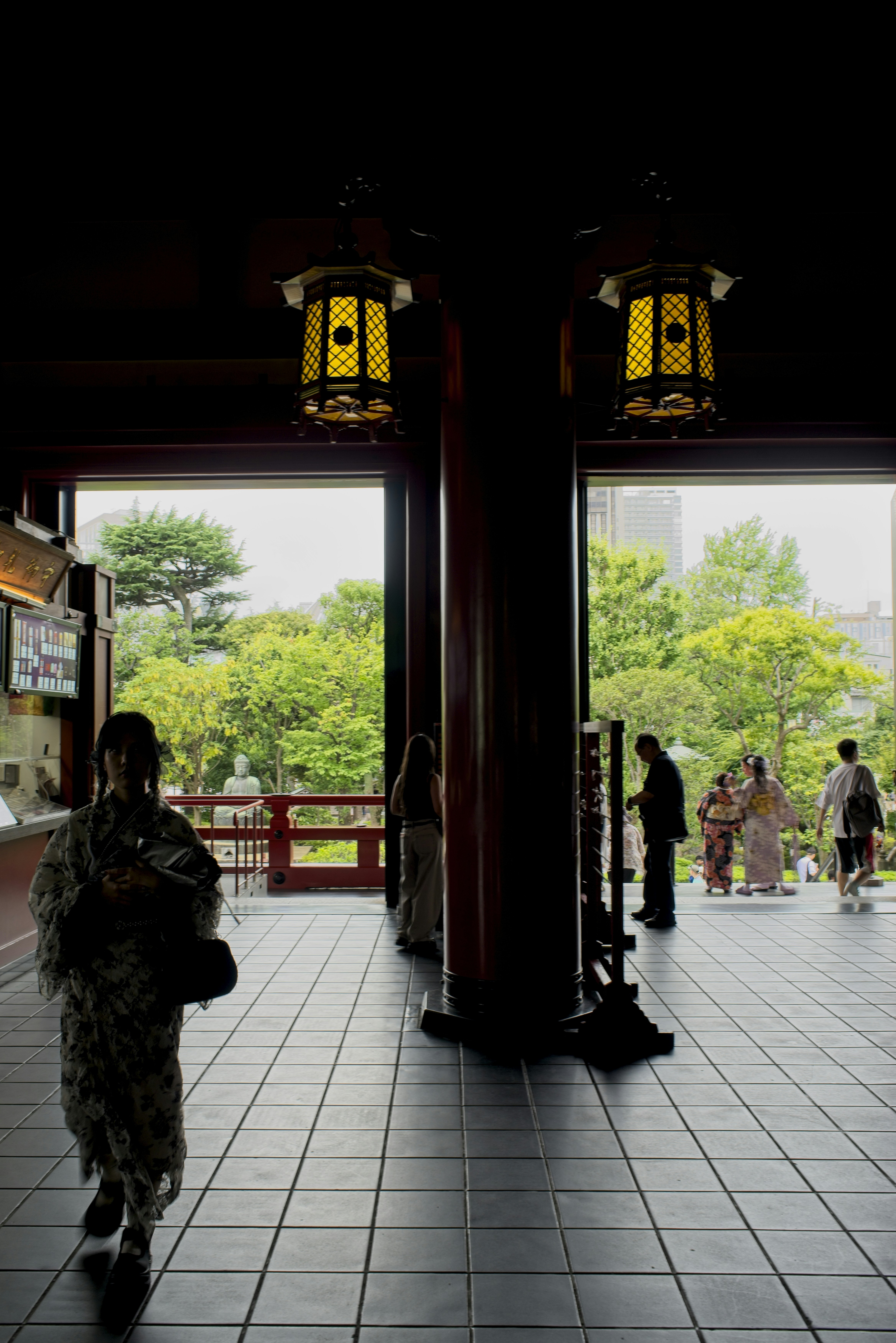 A woman in a floral kimono stands silhouetted near the entrance of Senso-ji's main hall, framed by glowing yellow lanterns hanging from the dark wooden ceiling. Beyond the temple's red pillars, visitors gather near a bronze Buddha statue visible through the open doorway, surrounded by lush greenery. The interplay of deep shadow and bright daylight creates a striking, atmospheric composition, capturing the quiet transition between sacred interior space and the temple's sunlit courtyard.