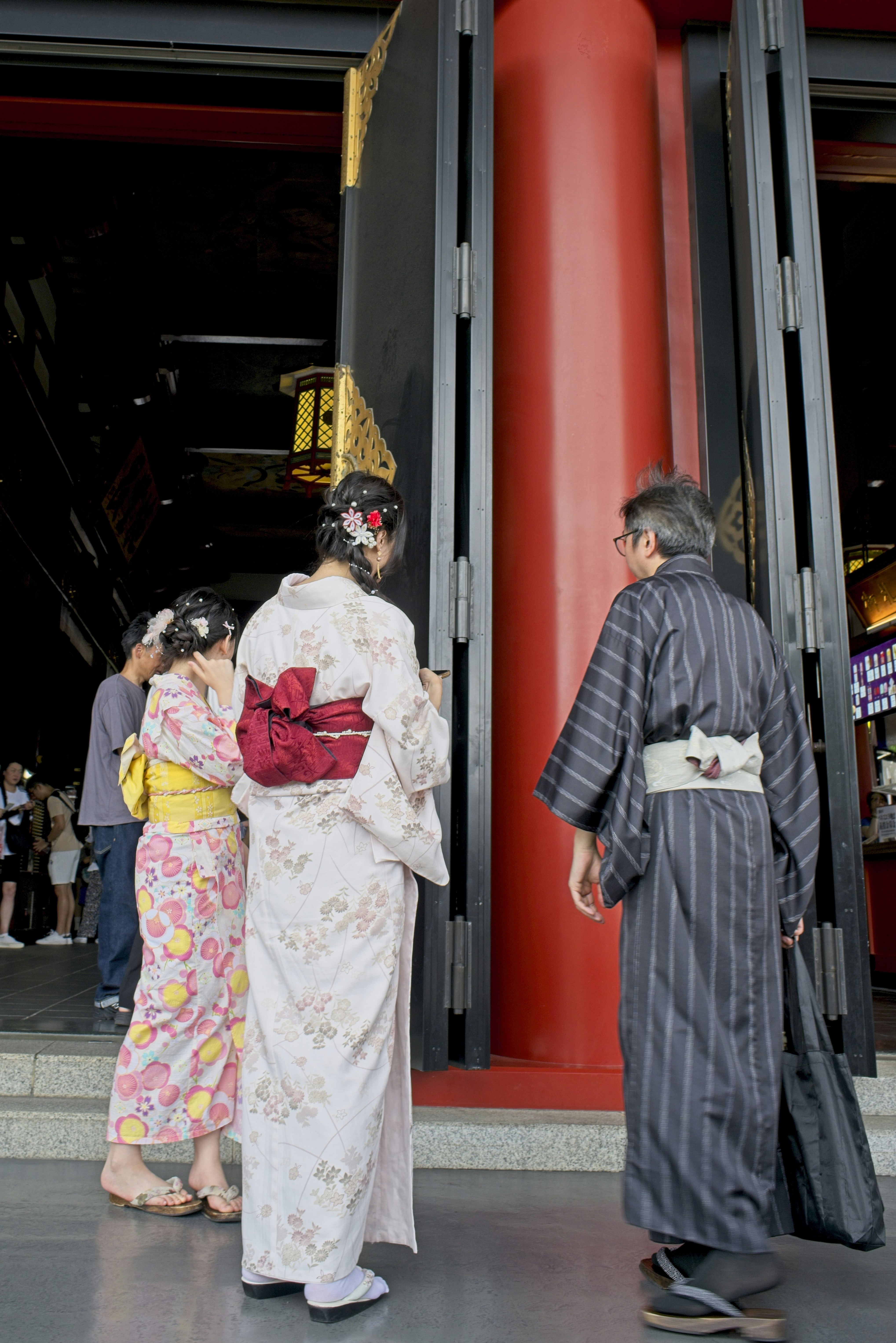 Three visitors dressed in traditional kimonos pause at the entrance of a Senso-ji temple gate, their elaborate obi sashes and floral hair ornaments adding vibrant detail against the striking red pillar. One woman wears a soft pink floral kimono with a bold crimson bow, while a man in dark pinstriped robes stands nearby. This candid moment captures the timeless elegance of Japanese traditional dress against Asakusa's iconic temple architecture on a warm July afternoon.