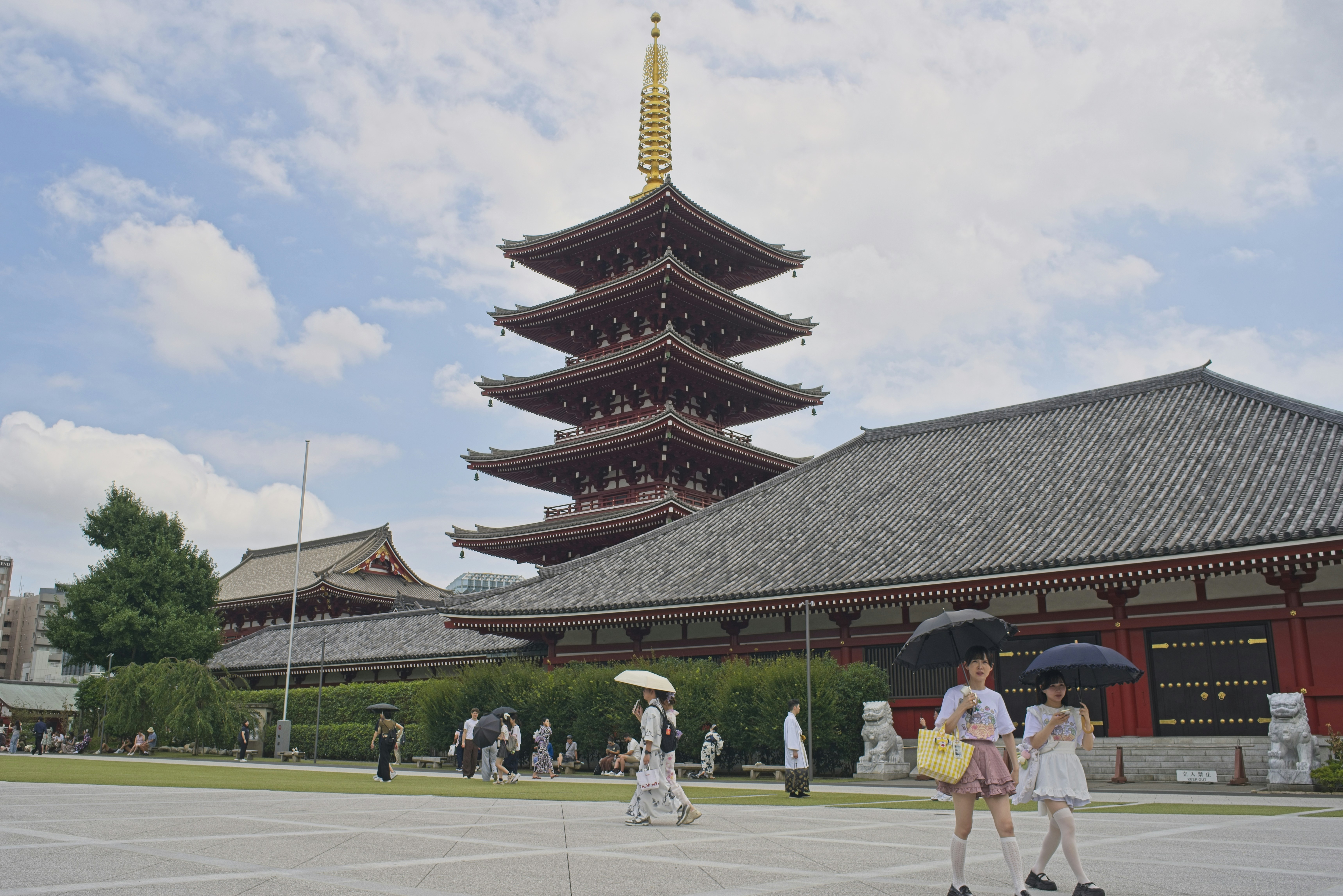 Senso-ji's Five-Story Pagoda rises majestically against a partly cloudy blue sky, framed by the temple's sweeping tiled roofline and a guardian stone lion statue. Visitors carry umbrellas for sun protection while strolling across the temple courtyard, including a woman in a traditional kimono and two friends dressed in playful street fashion. This vibrant scene captures the timeless architectural grandeur of Asakusa alongside contemporary Tokyo style on a warm July afternoon.