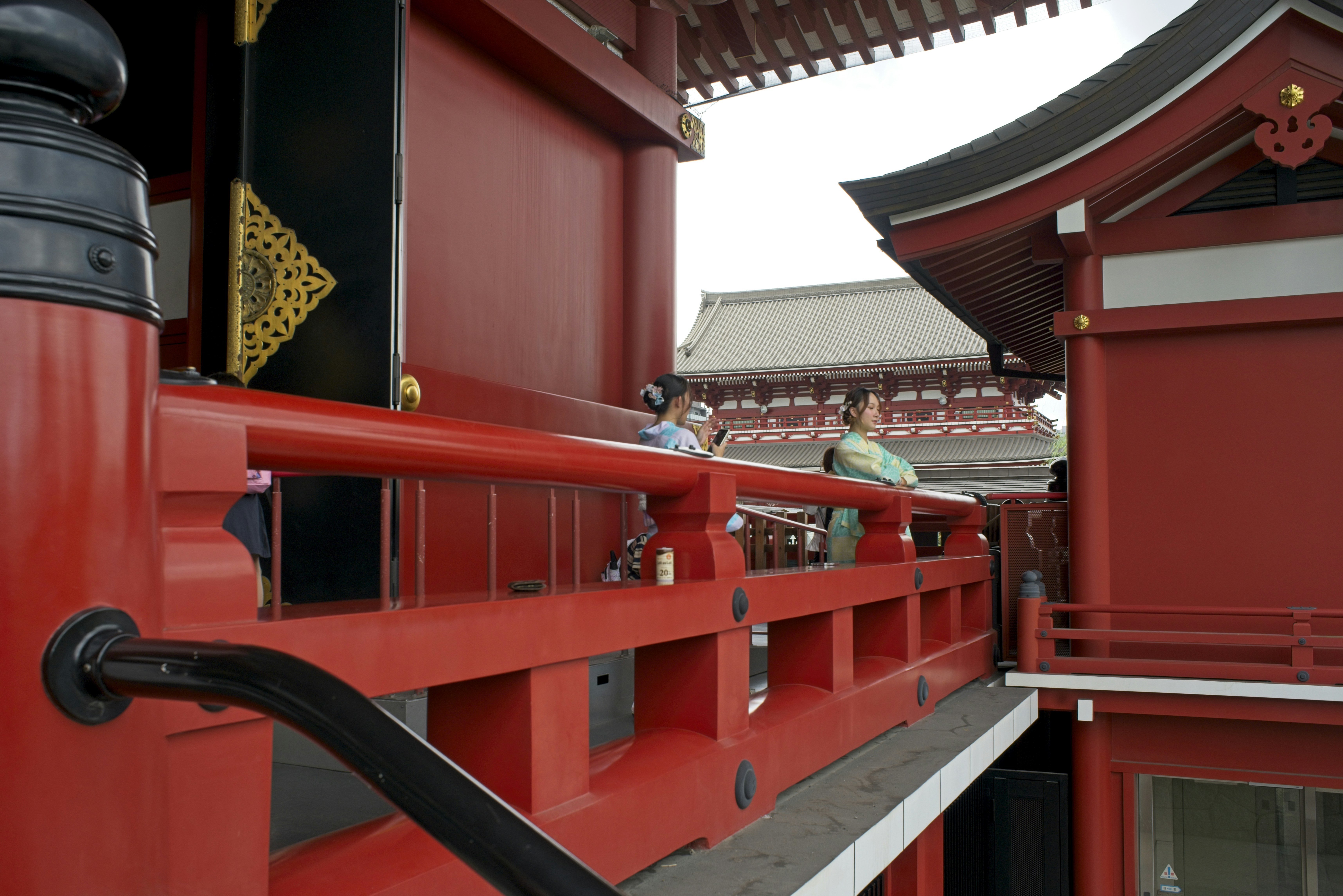 Two visitors in traditional kimonos lean against the vivid red railing of a temple balcony at Senso-ji, framed by bold architectural details including gold floral motifs and layered wooden beams. The tiered rooflines of neighboring temple structures rise in the background against a hazy sky. This close-up architectural composition highlights the intricate craftsmanship and vibrant color palette that define Senso-ji's iconic buildings, capturing a quiet moment of contemplation amid Asakusa's historic temple grounds.