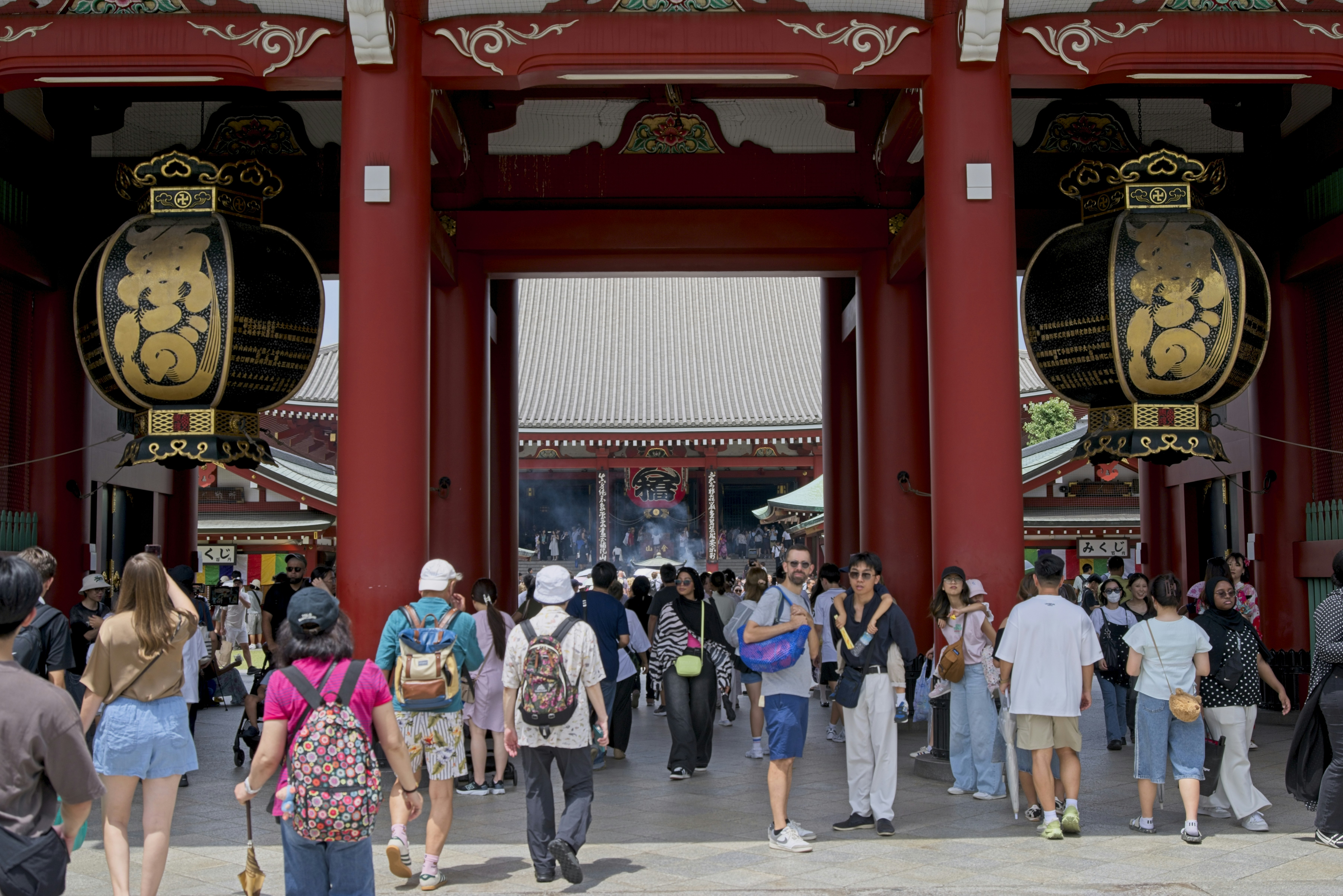 A dense crowd of tourists passes beneath Senso-ji's Hozomon Gate, flanked by massive black-and-gold lanterns hanging from the towering red pillars. Beyond the gate, the temple's main hall comes into view, its incense smoke rising near the entrance steps where visitors gather. Backpacks, sun hats, and casual summer attire fill the frame as travelers navigate the busy walkway. This vibrant scene captures the constant flow of foot traffic through one of Tokyo's most iconic temple entrances on a hot July afternoon.