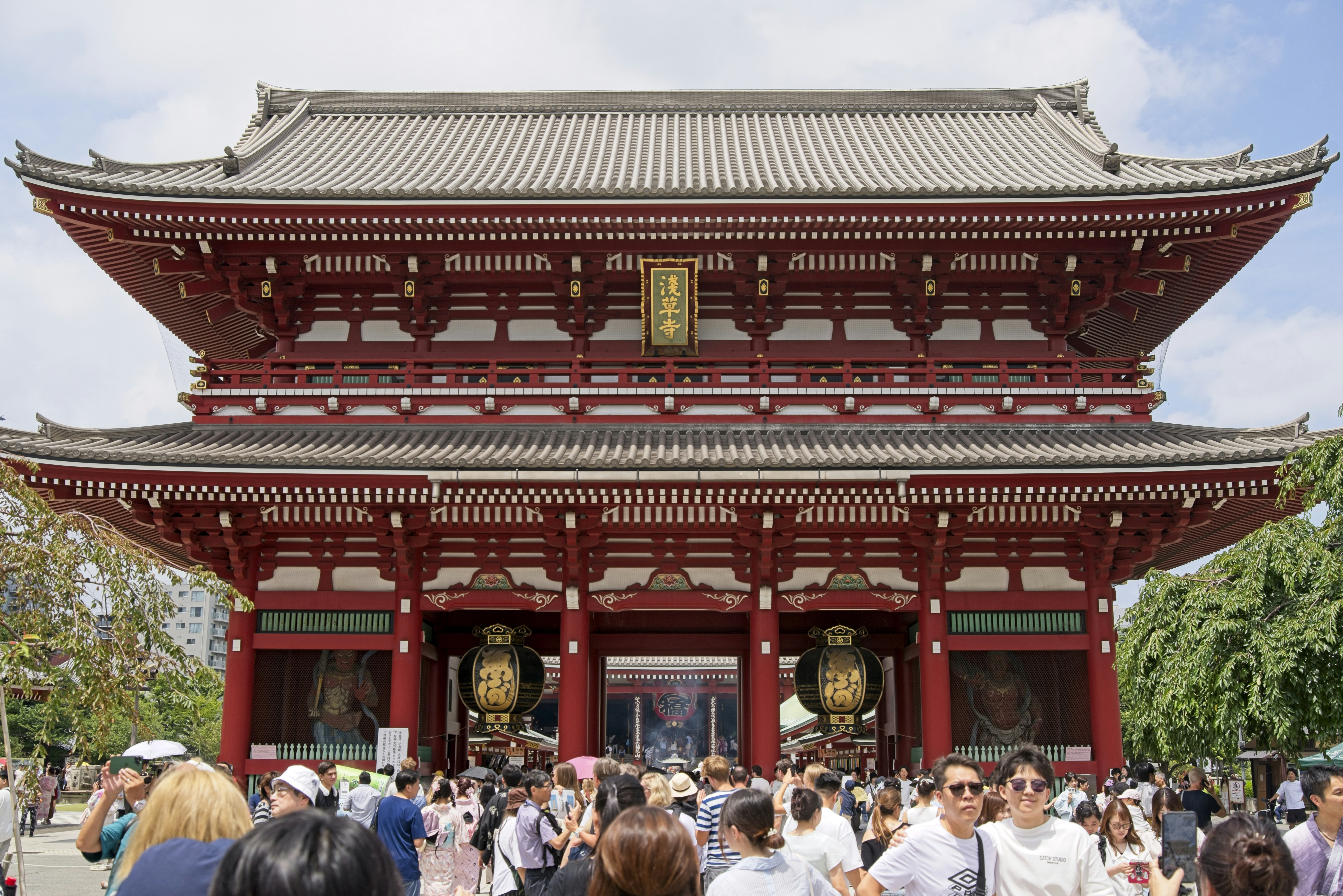 Senso-ji's grand Hozomon Gate towers above a dense crowd of visitors, its striking red pillars and tiered grey roof framing two massive black-and-gold lanterns hanging beneath the entrance. Guardian statues stand watch on either side of the doorway, while a golden nameplate reading "Sensoji" adorns the upper level. Tourists and locals gather beneath the gate, capturing photos and soaking in the temple's grandeur. This iconic entrance showcases the timeless architectural beauty of one of Tokyo's most beloved religious sites.