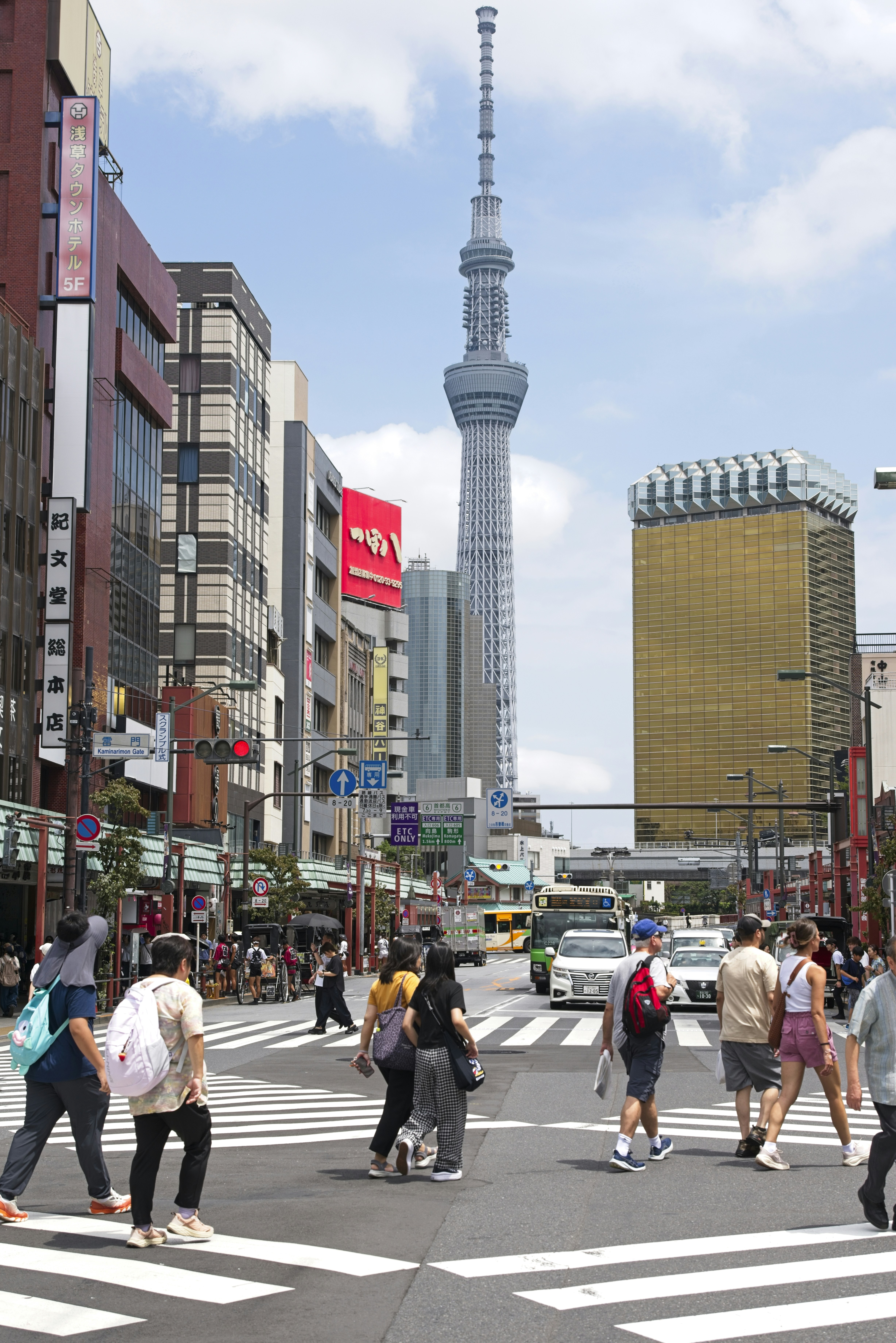 Tokyo Skytree pierces the sky above Asakusa's busy streets, framed perfectly between rows of buildings as pedestrians cross a wide intersection near Kaminarimon Gate. Buses, cars, and delivery trucks navigate the road below, while shoppers and tourists carrying bags and backpacks move through the crosswalk. Colorful storefront signage lines both sides of the street, blending traditional Asakusa charm with the modern silhouette of Japan's tallest tower rising dramatically in the background under a bright July sky.