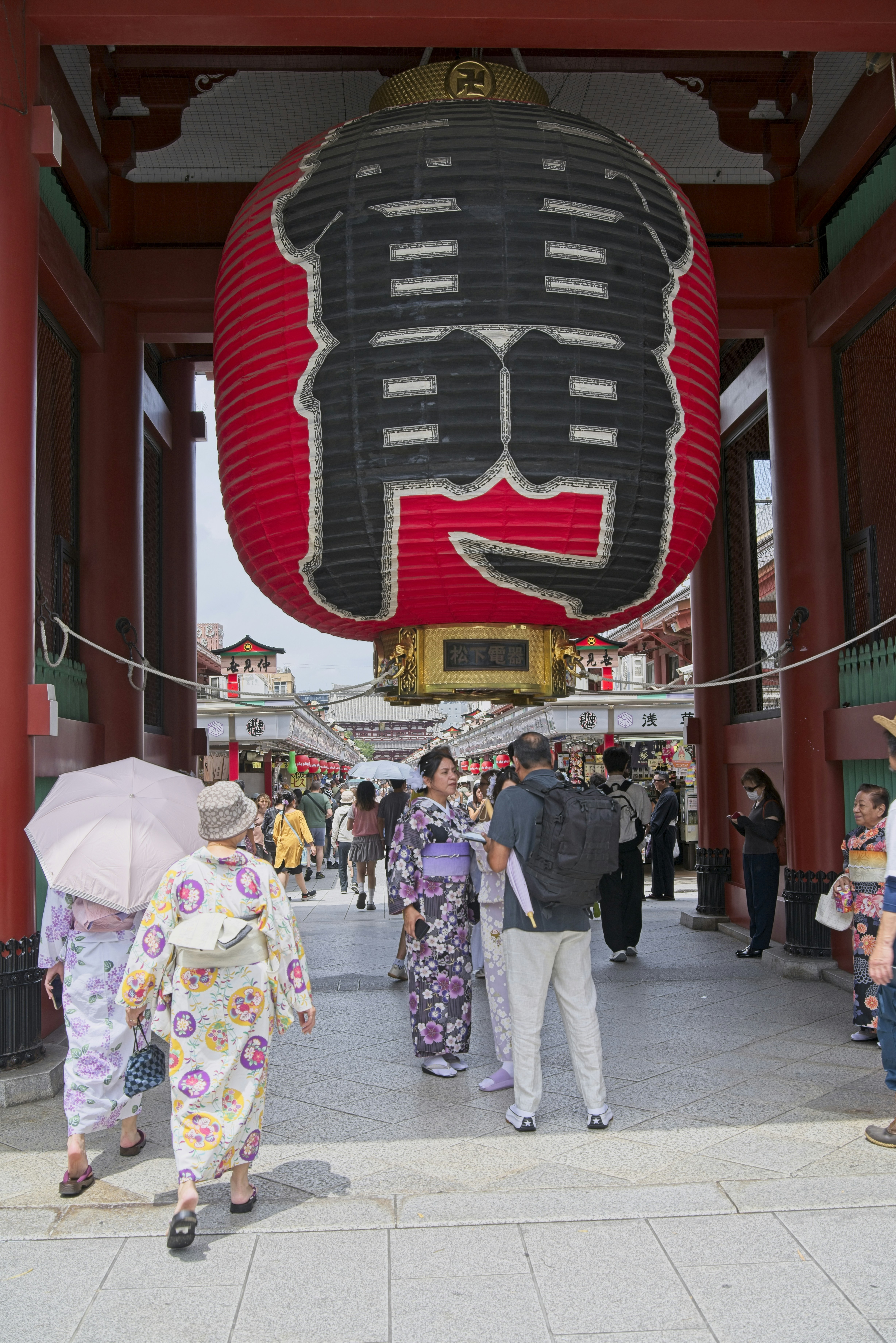 The massive red-and-black lantern of Kaminarimon, Senso-ji's iconic Thunder Gate, dominates this view down Asakusa's bustling Nakamise shopping street. Visitors in traditional kimonos mingle with tourists carrying umbrellas for sun protection, all passing beneath the towering paper lantern inscribed with bold Japanese characters. Shops and additional gates line the corridor stretching into the distance. This quintessential Asakusa scene captures the timeless grandeur of one of Tokyo's most photographed landmarks on a busy July afternoon.