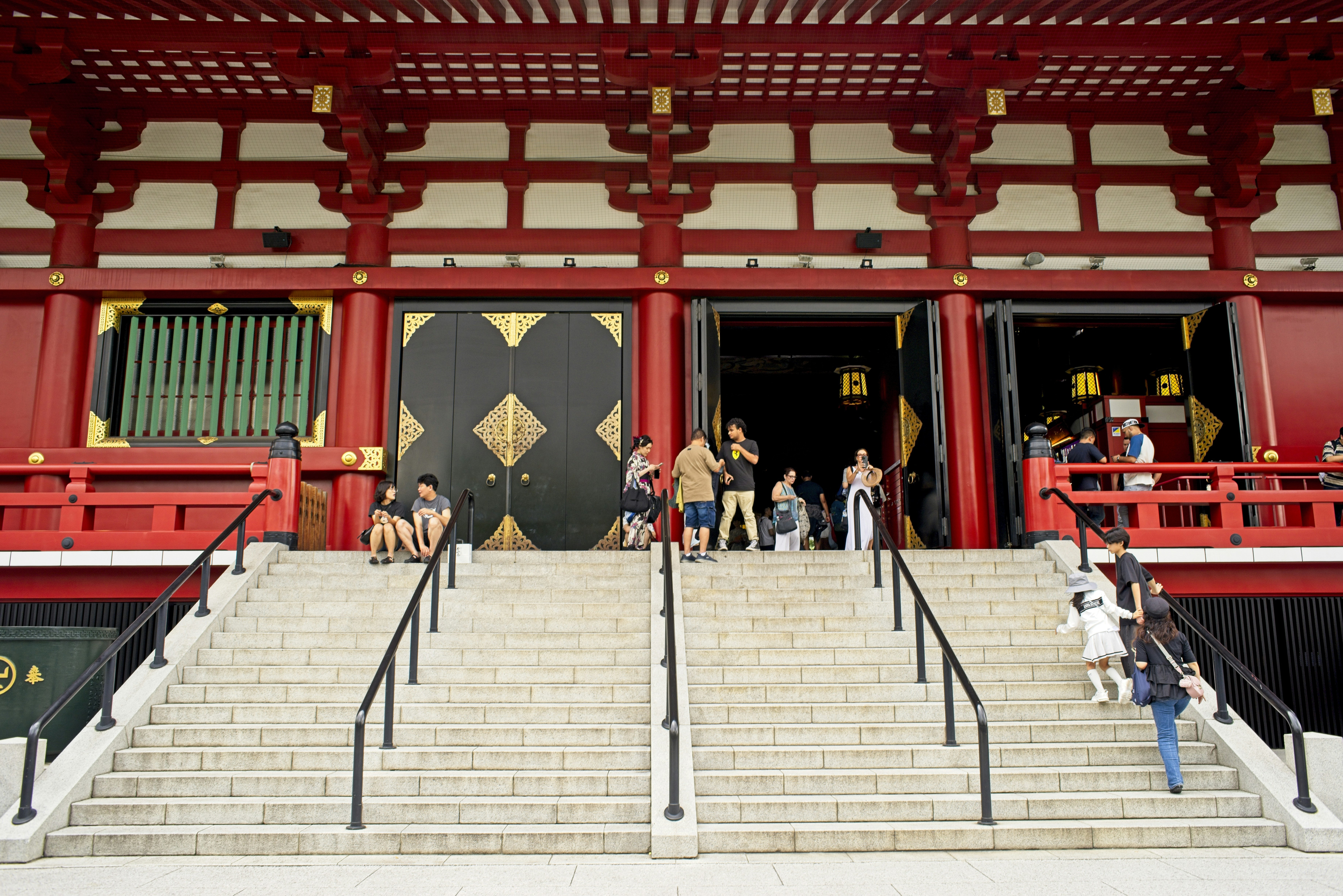 Visitors ascend and descend the wide stone steps leading to Senso-ji's main hall, its striking red pillars and gold-accented black doors framing the entrance. A woman in traditional kimono stands among casually dressed tourists near the open doorway, where glowing lanterns hint at the temple's interior. A family holds hands while walking down the stairs in the foreground. This candid scene captures the timeless architectural grandeur and everyday foot traffic of one of Tokyo's most iconic religious sites.