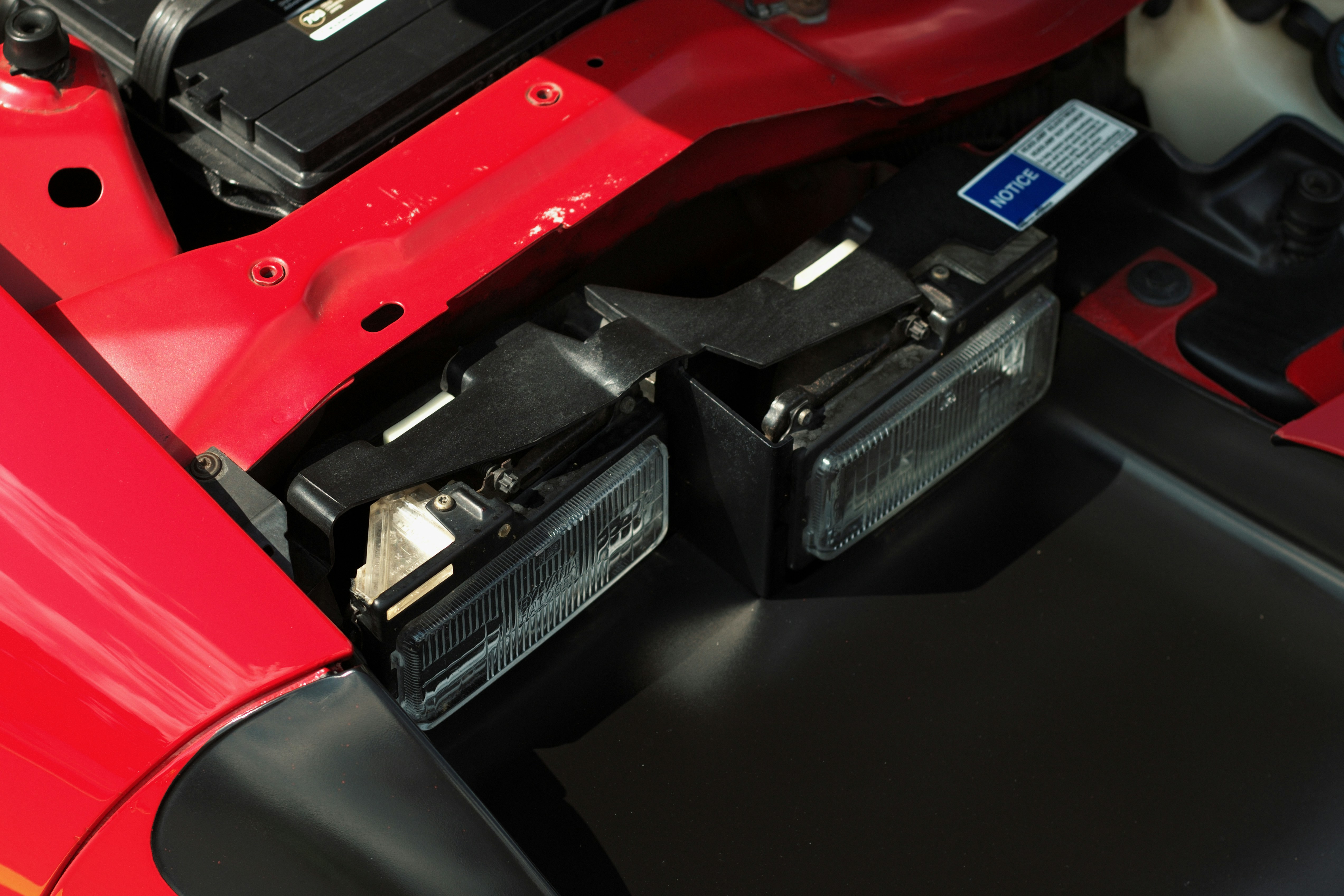 Front-angled view beneath the hood of a red sports car, exposing twin headlights and battery bay