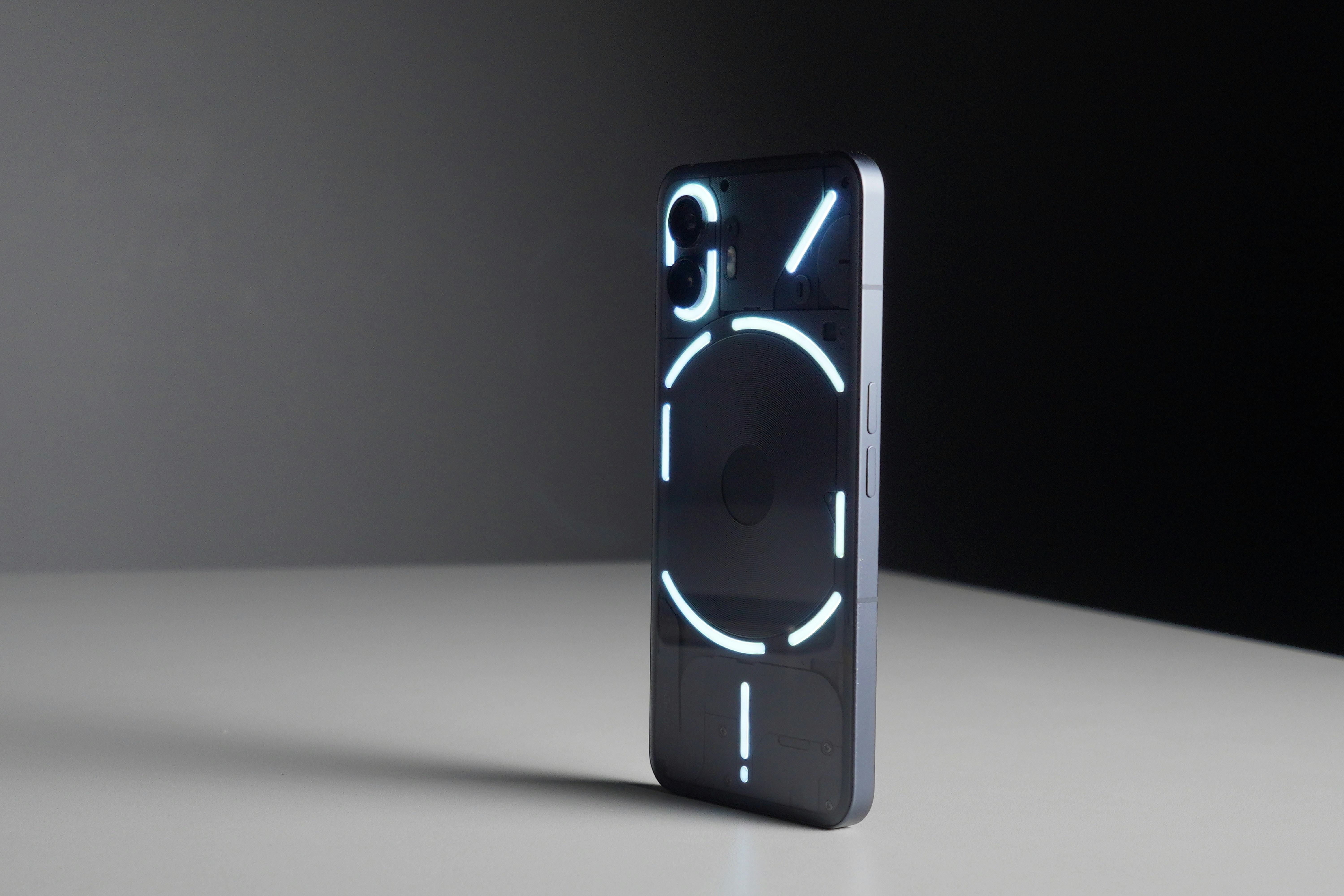 A sleek smartphone stands upright on a smooth surface, its back glowing with geometric neon lines. The minimal setup emphasizes the device as the focal subject with a shallow, moody backdrop.