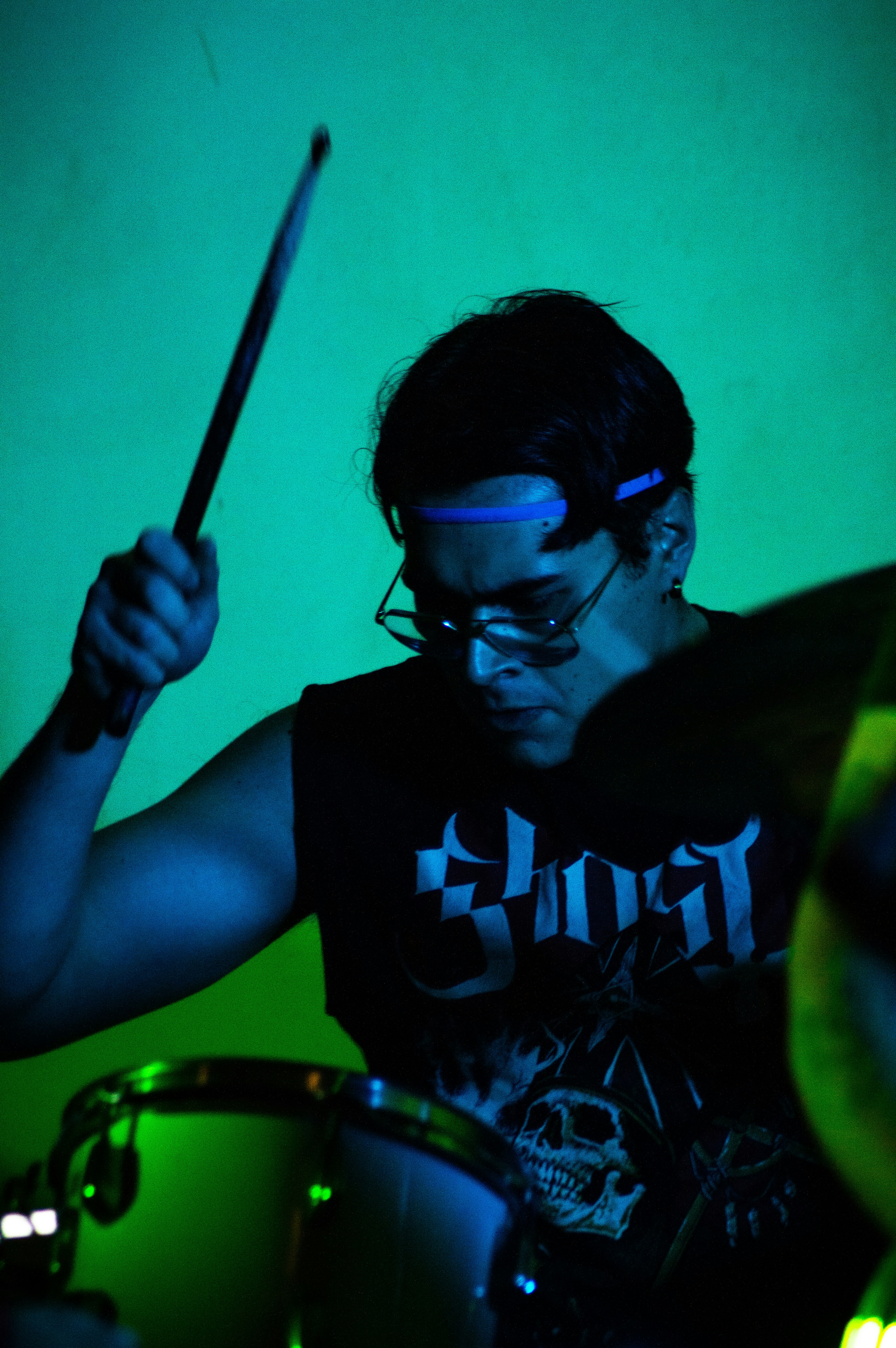 Drummer immersed in neon-green/blue stage lighting, with drumsticks raised and intensity focused on the kit; a bold graphic shirt and glasses catch the spotlight.