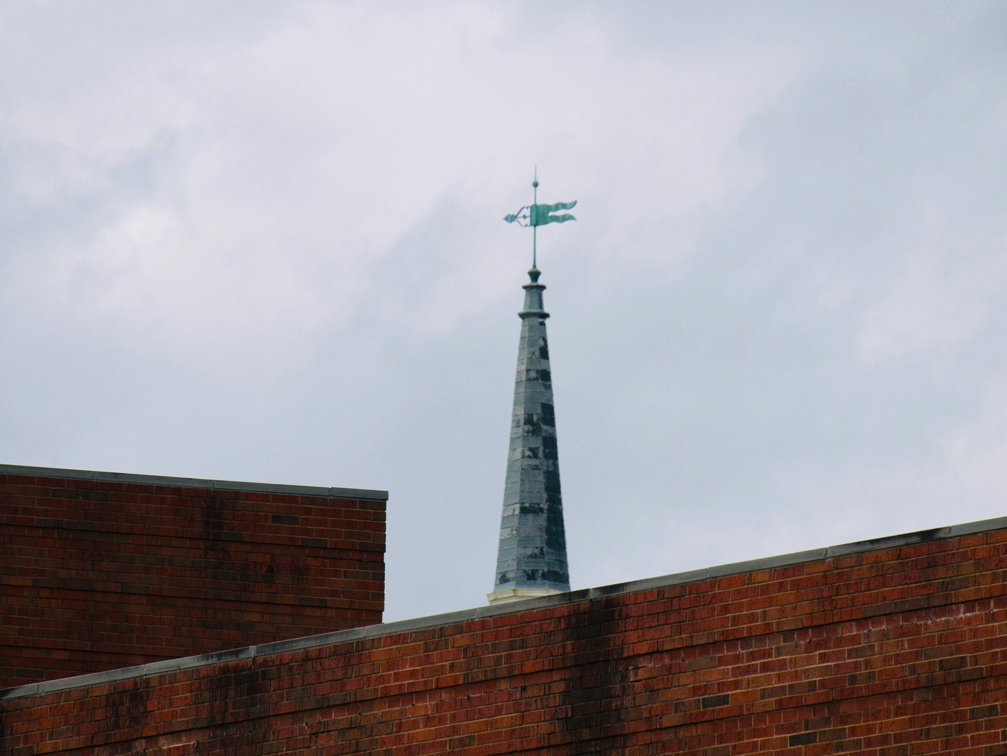Red-brick rooftop with a weather vane-topped steeple piercing a pale sky, the vertical spire rising behind slanted rooflines. A minimal architectural scene without people.