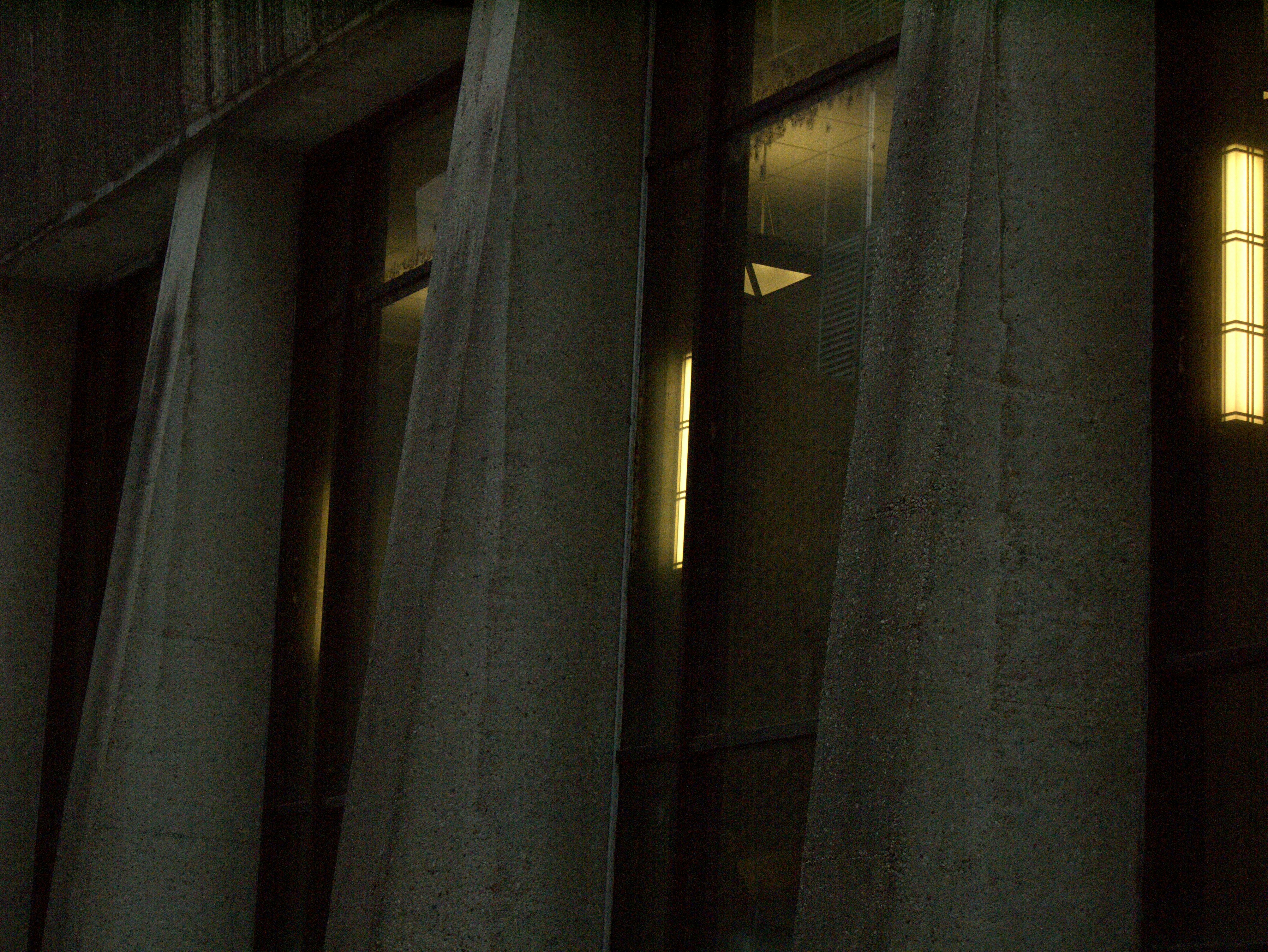 Muted architectural detail with towering concrete columns and narrow vertical windows glowing softly, suggesting an urban interior or exterior sculpture. A study in texture and light.