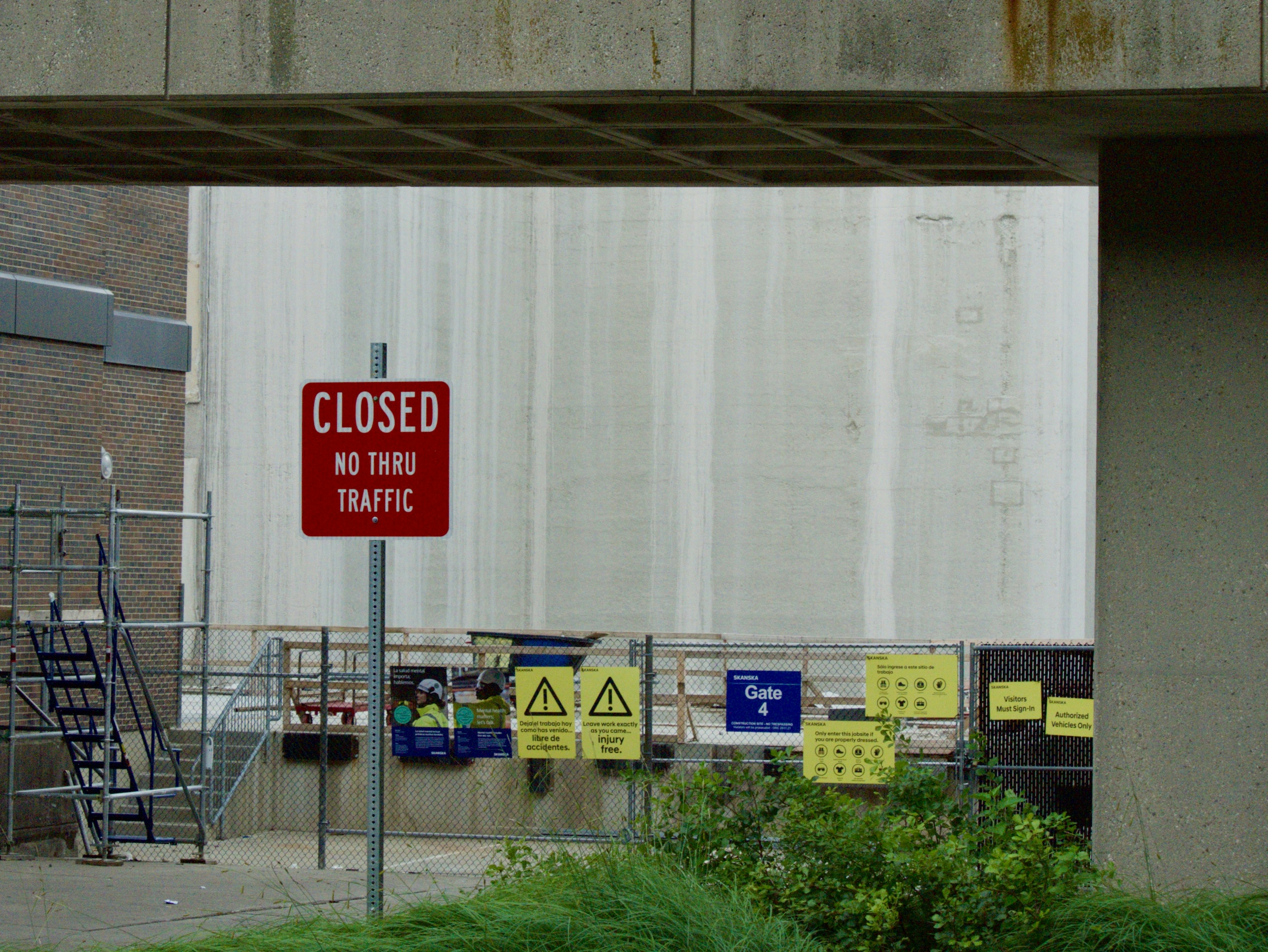 A construction site behind a chain-link fence with multiple yellow warning signs and a prominent red 'Closed' sign under an overpass.