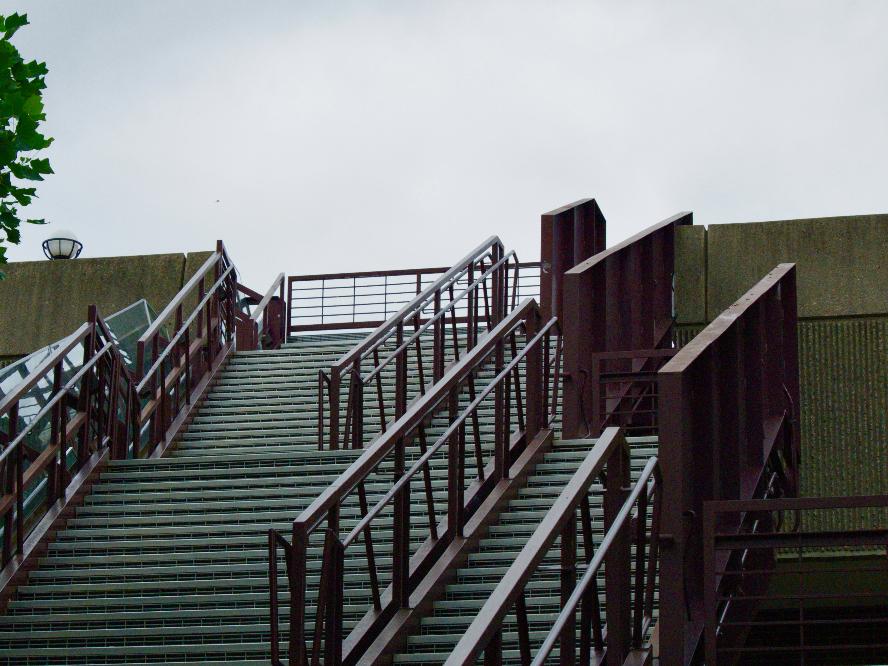 Industrial staircase climbing toward a pale sky, steel rails and geometric forms create a dramatic ascent. Photograph highlighting architectural lines and scale.