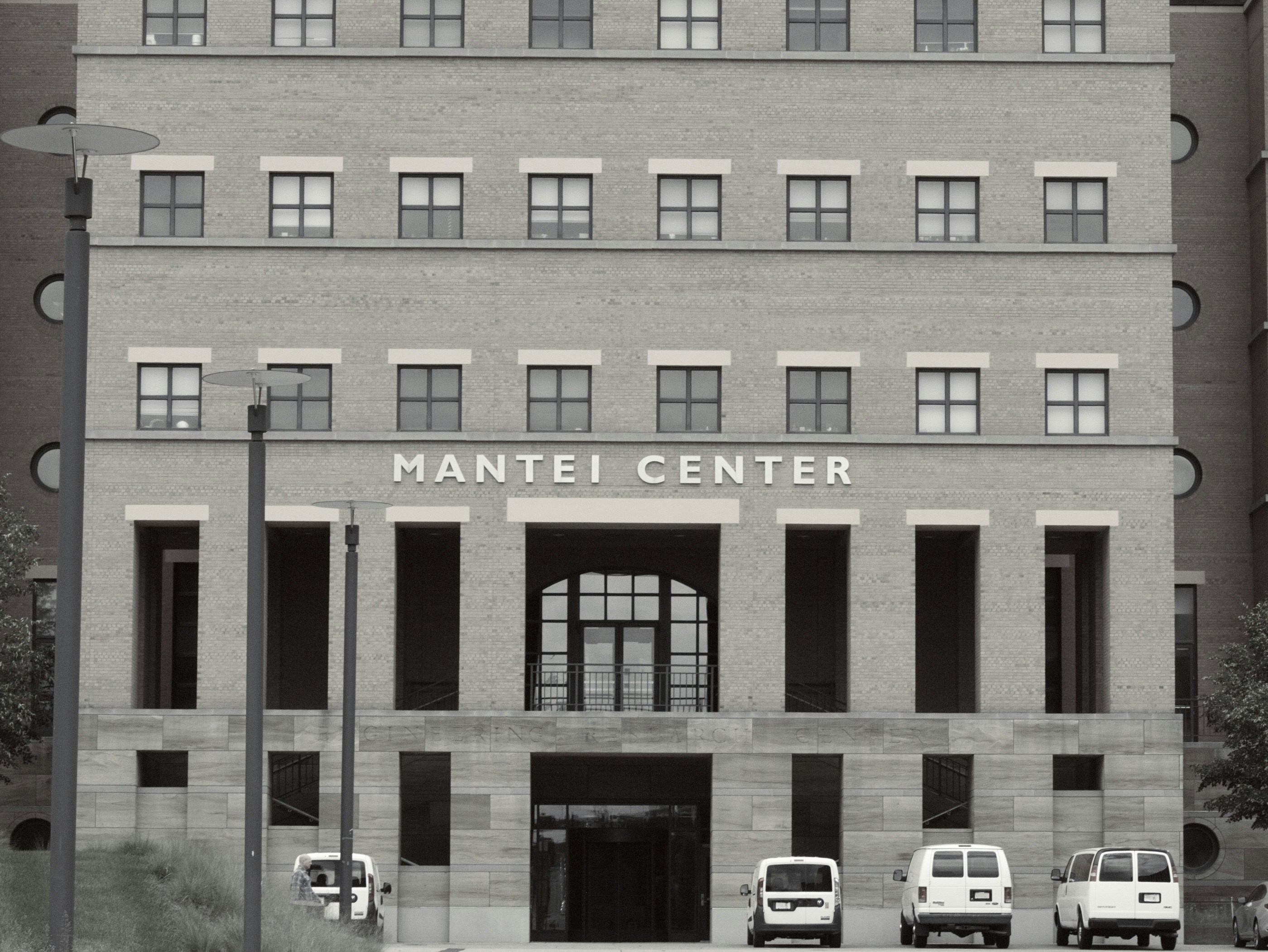 Symmetrical brick facade of the MANTEL Center with arched entry and parked cars along the base, captured in grayscale to emphasize architectural detail.