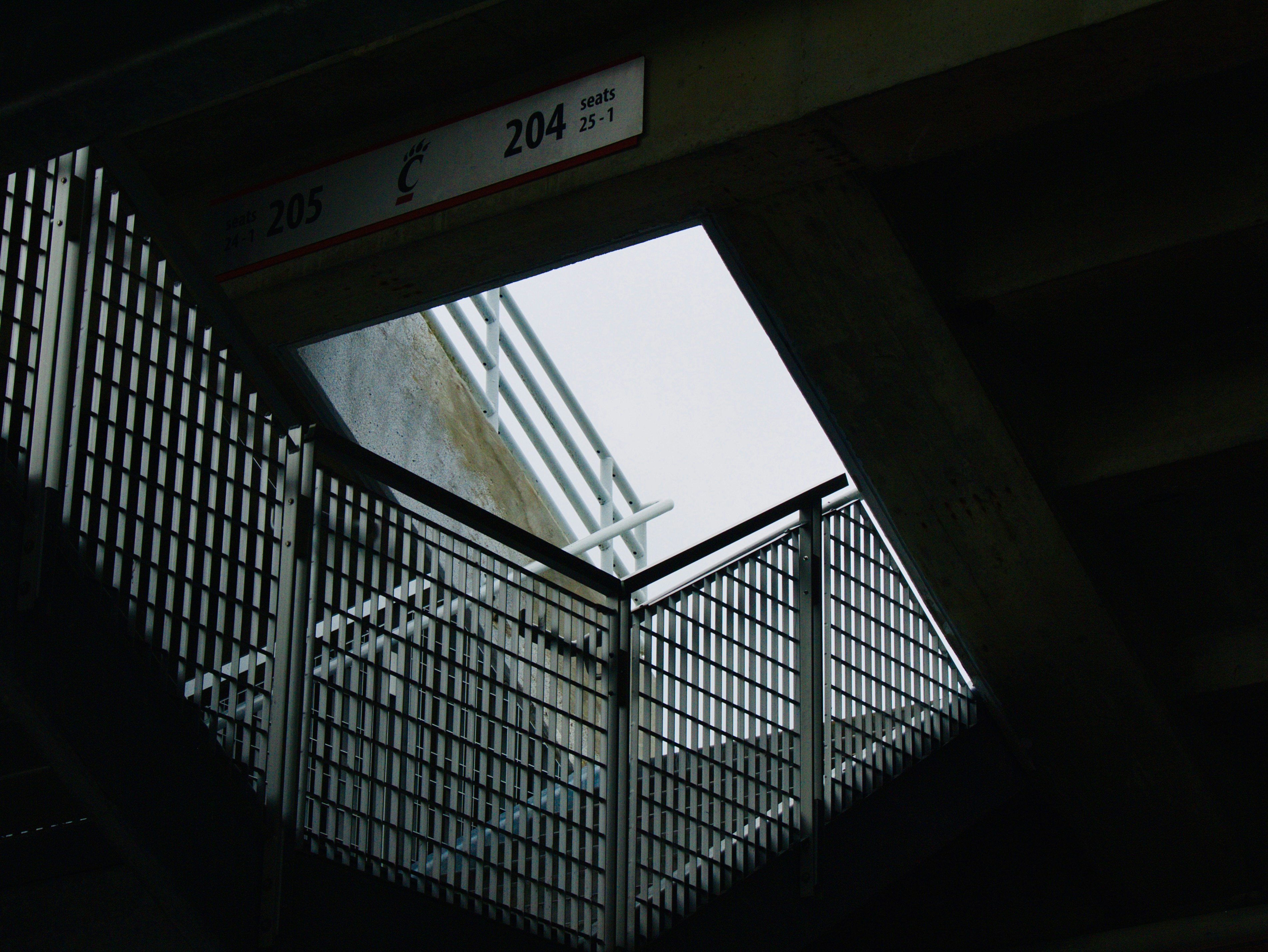 Angular stairwell fencing and concrete beams silhouette a pale sky, captured through a geometric opening in a dim structure. The scene emphasizes architectural lines and negative space.