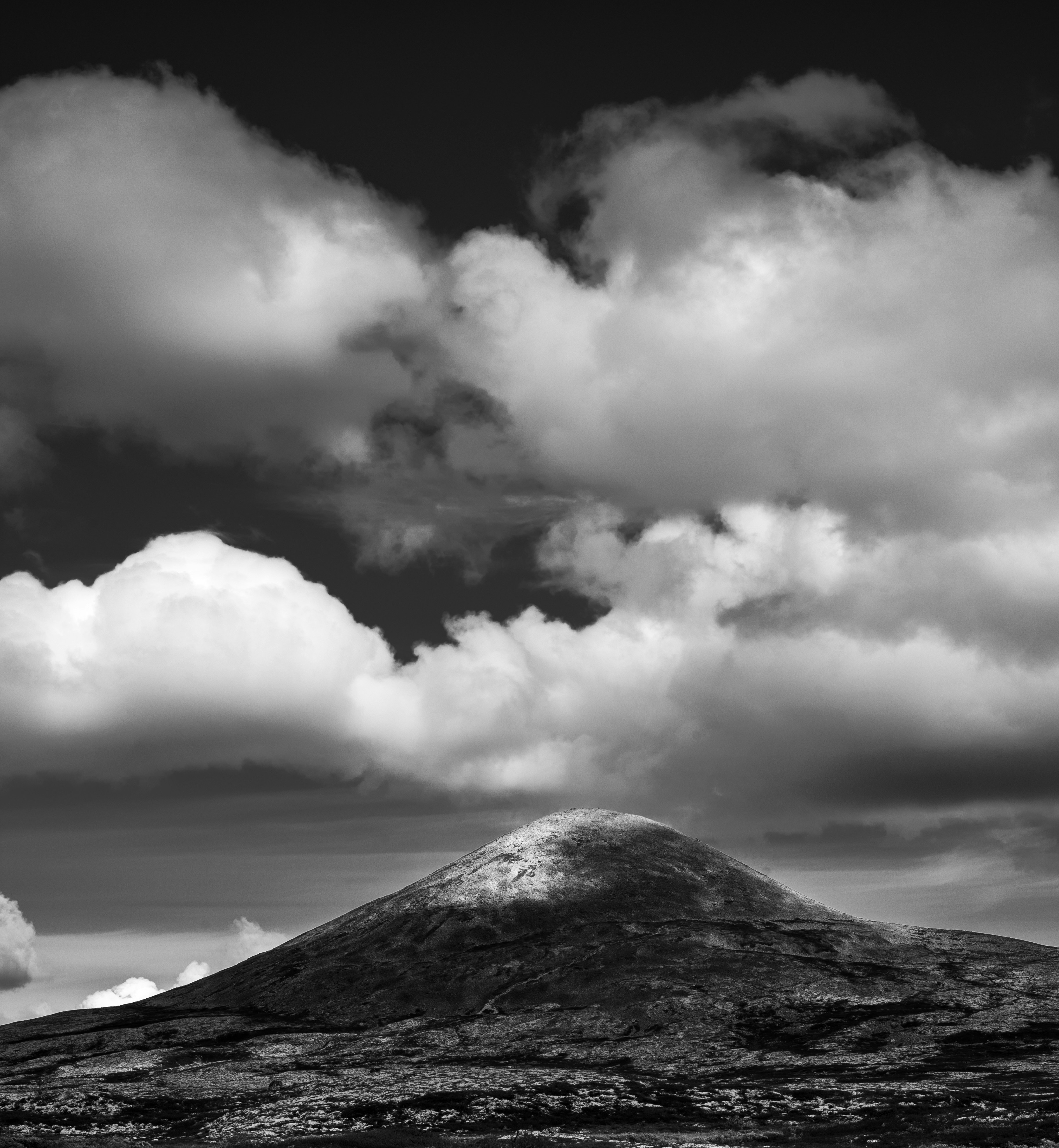 Monochrome landscape featuring a solitary, rounded hill beneath dramatic clouds, emphasizing texture and contrast in the terrain.