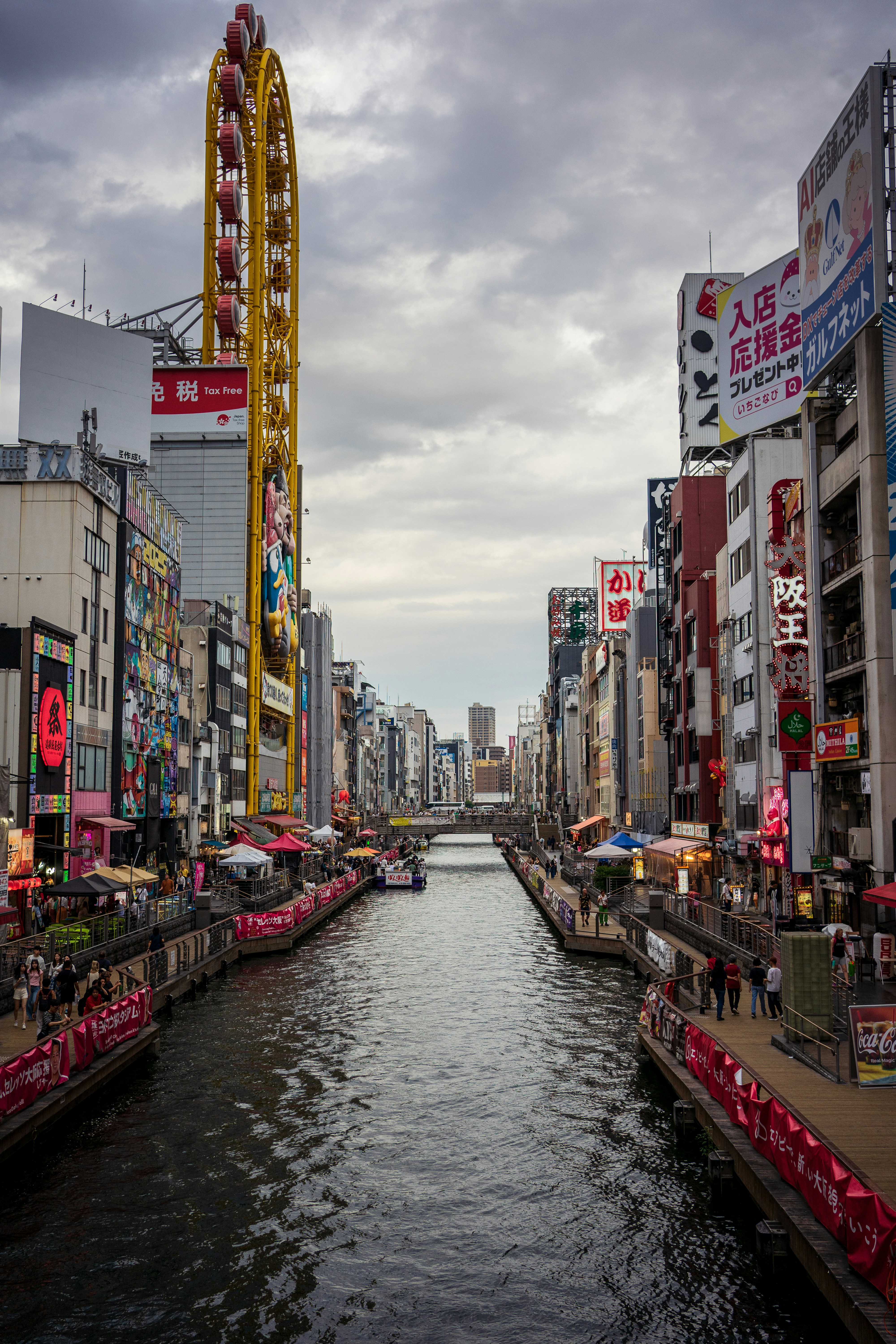 Bustling canal bisects a vibrant urban arcade flanked by towering signage and a towering Ferris wheel, capturing a lively riverside thoroughfare at dusk.