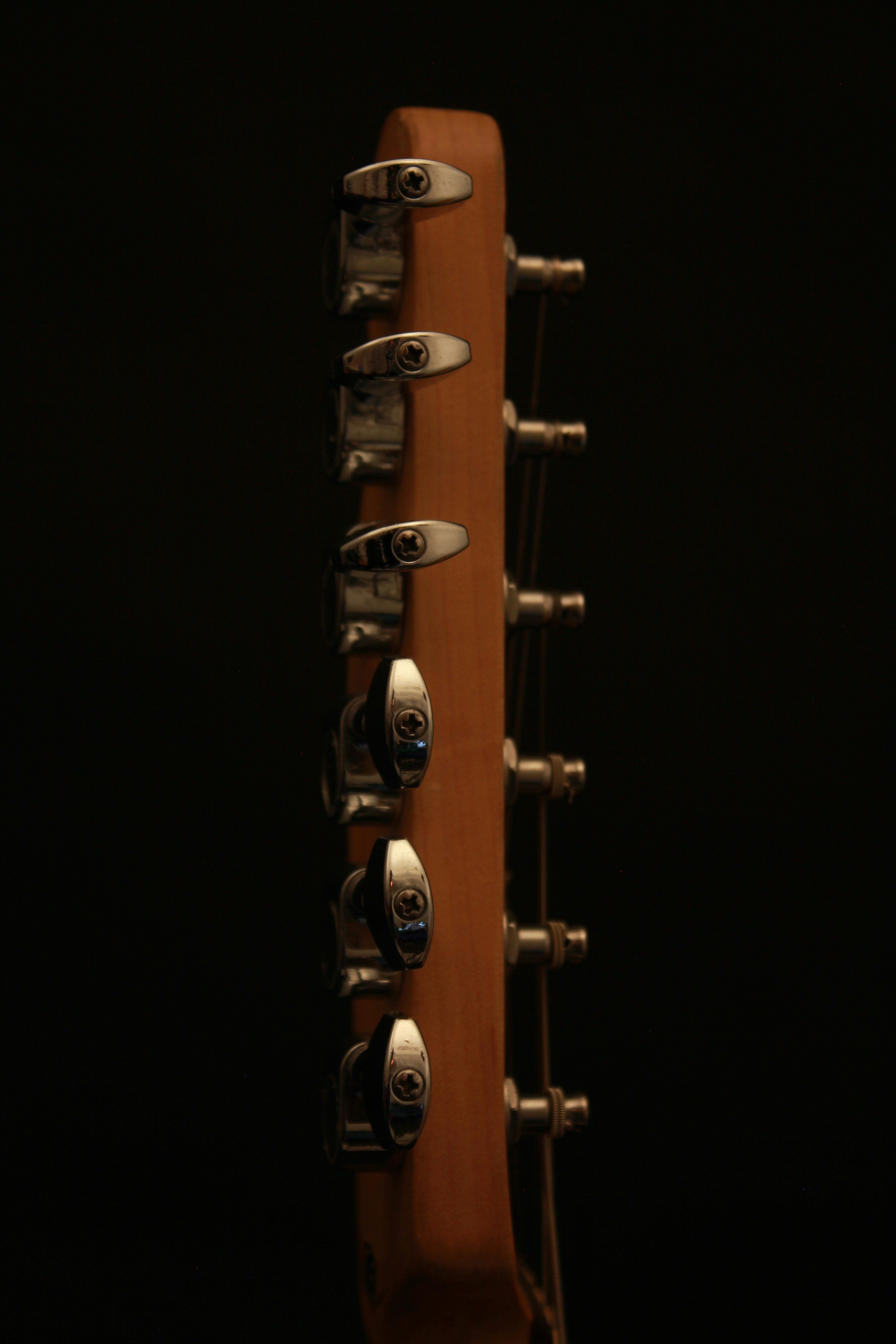 Close-up of a guitar headstock emerging from a dark background, highlighting tuning pegs and wood grain with subtle warm lighting.
