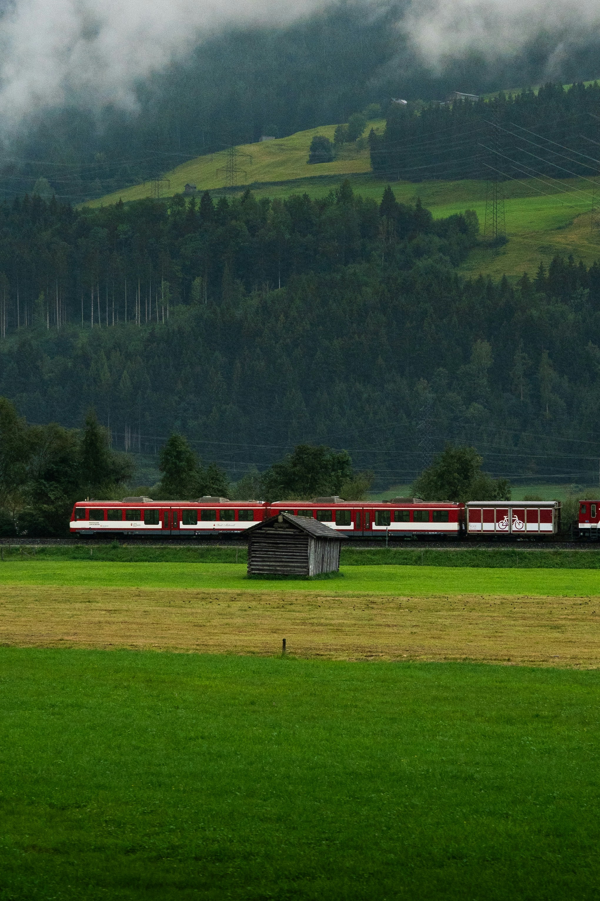 Long red-and-white passenger train traverses a flat rural landscape with a small wooden shed in the foreground and forested hills in the background.