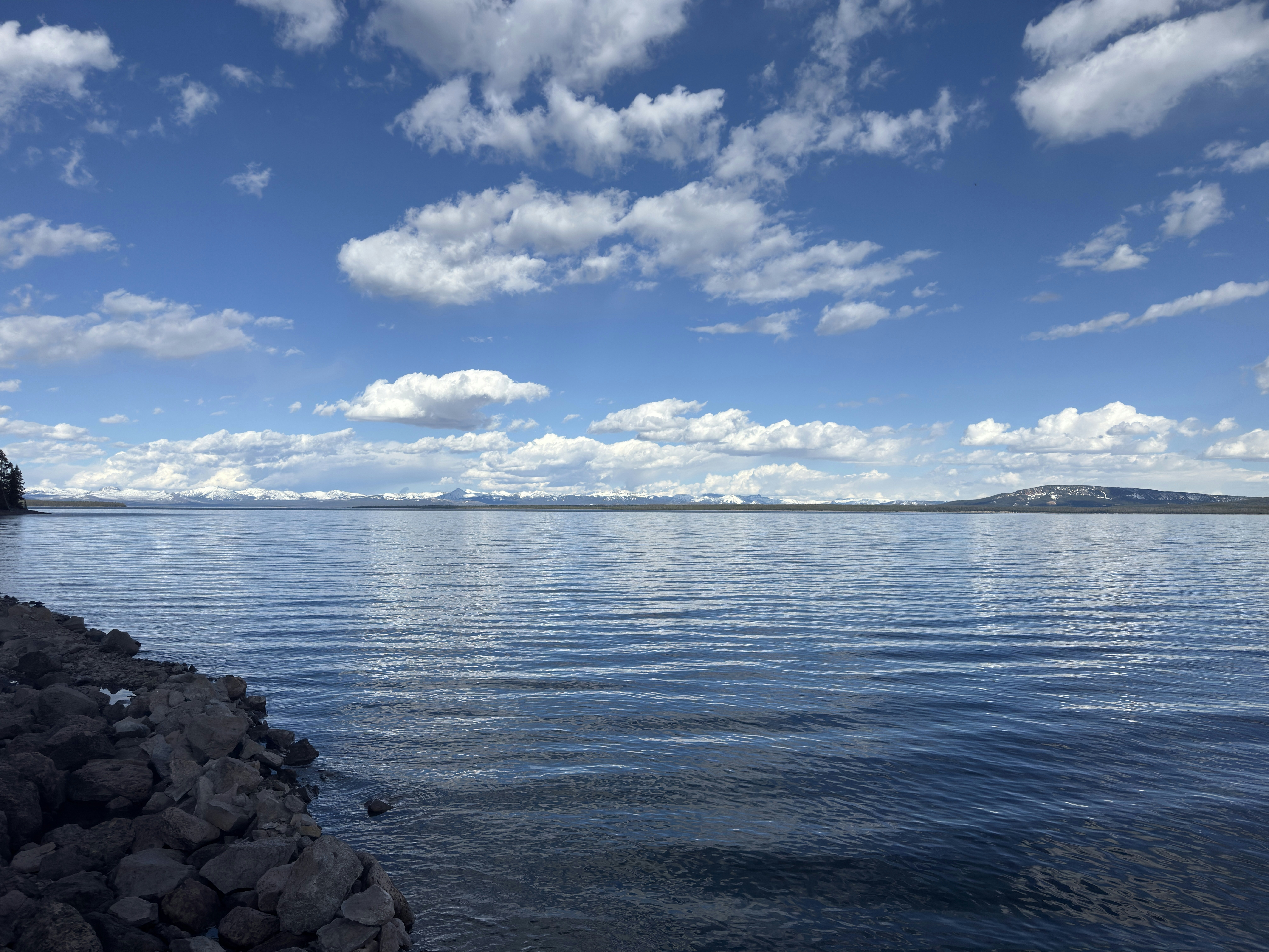 Clear lakeshore with rocky breakwater and calm water under a bright blue sky; a tranquil landscape emphasizing depth and reflection.