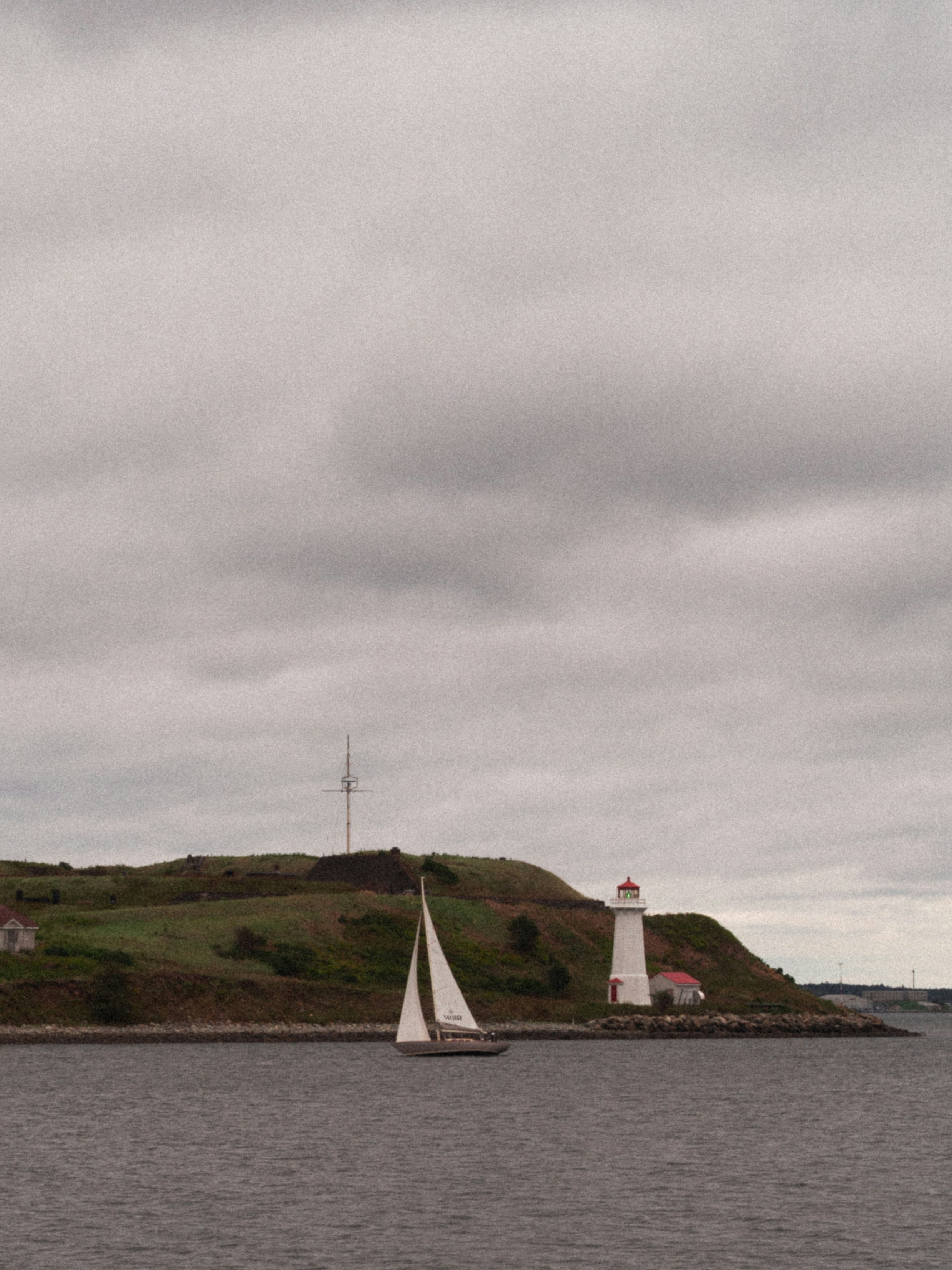 Sailboat glides past a coastal lighthouse on a calm day; muted light and overcast sky add atmosphere to the seascape.