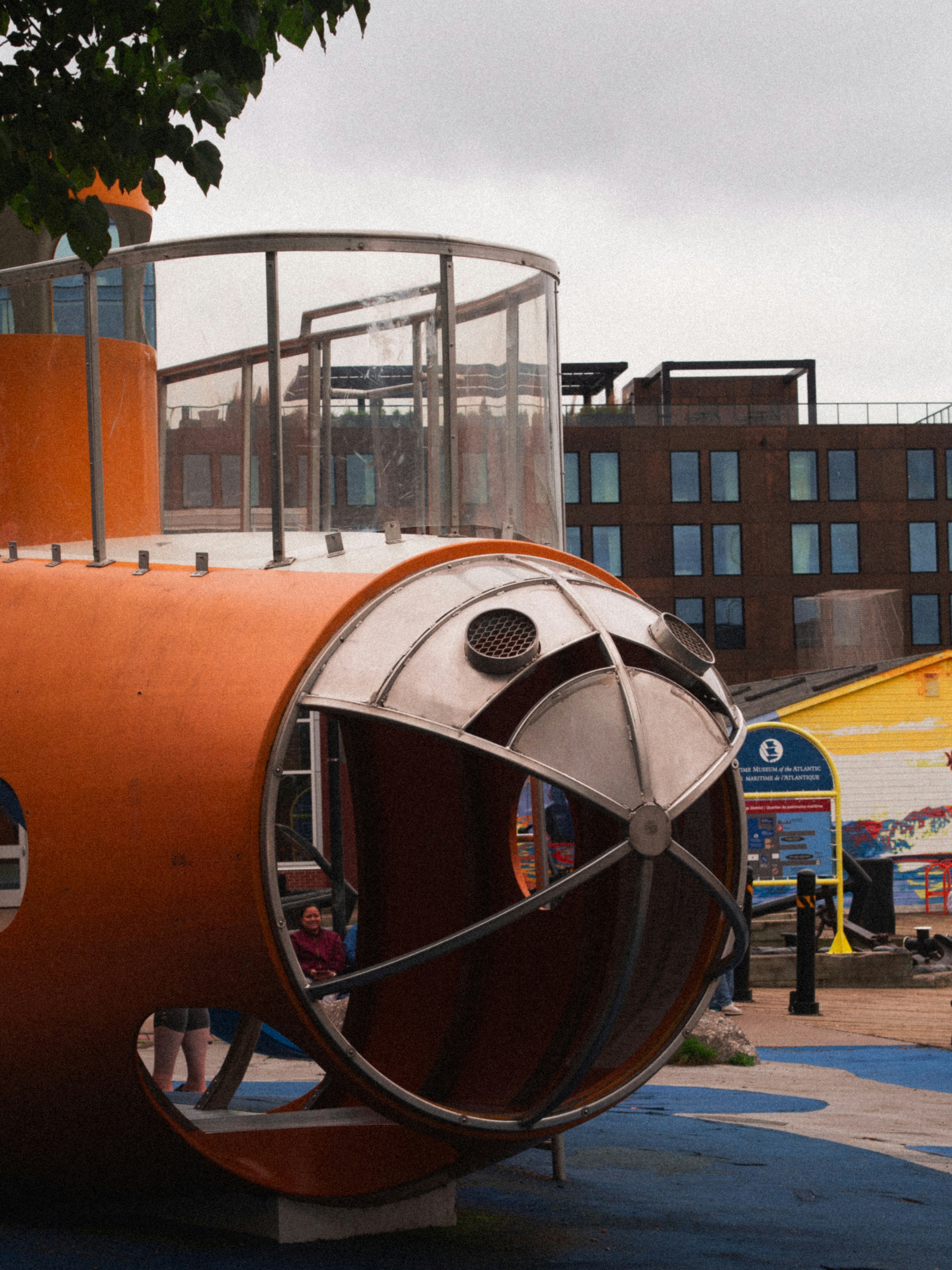 Orange playground tunnel with metallic dome sits in foreground; urban buildings and a yellow/blue mural visible in background.