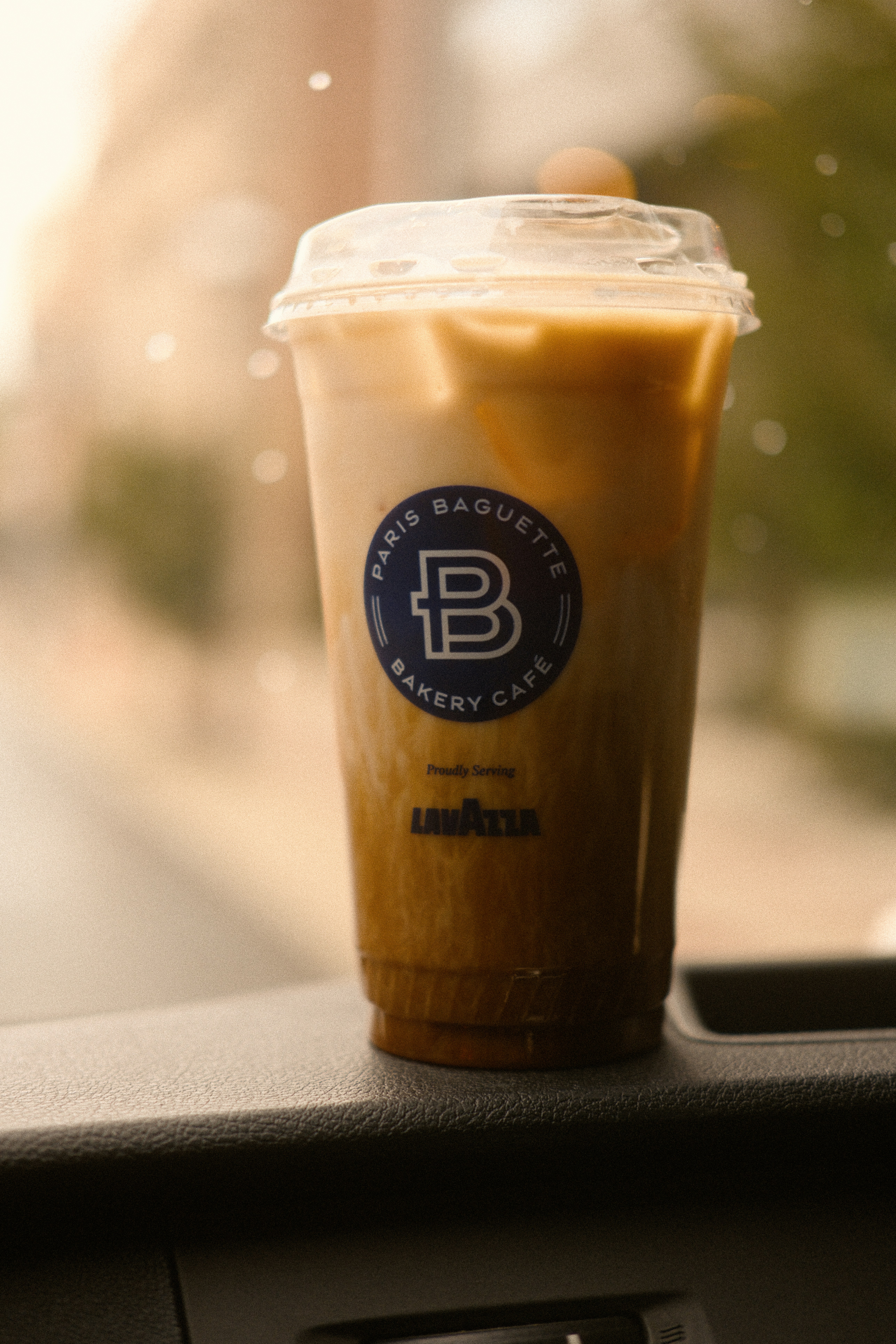 A tall iced latte from Paris Baguette sits on a car dashboard, blurred cityscape visible through the window. The logo on the cup is centered, with warm bokeh and condensation adding mood. Logo and branding indicate a café beverage.