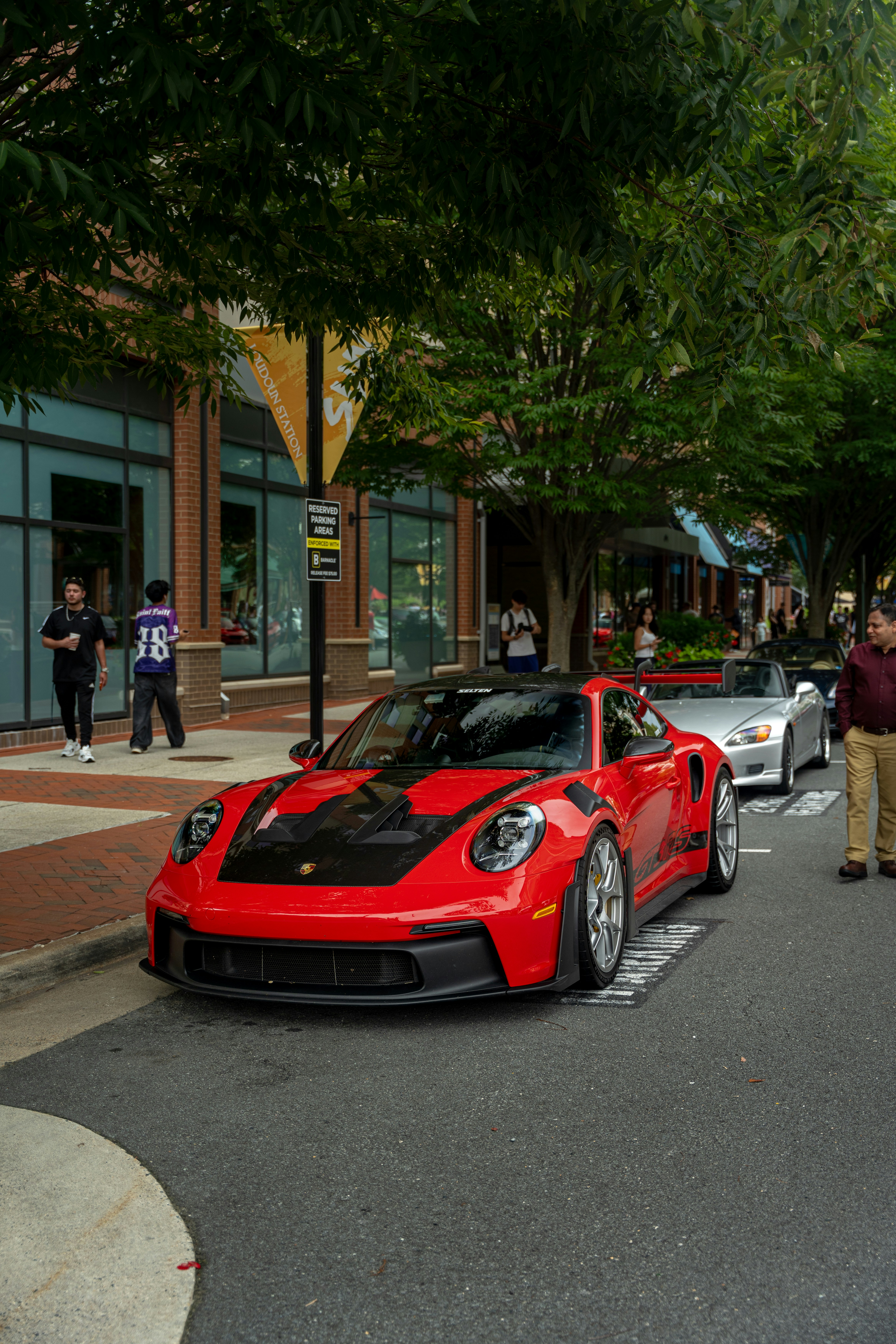 Red Porsche 911 GT3 RS parked on a city street with pedestrians and shops in the background. The scene captures a moment of urban automotive display with surrounding greenery and passersby. Car is clearly the focal subject, with the shot presenting ambient street context.