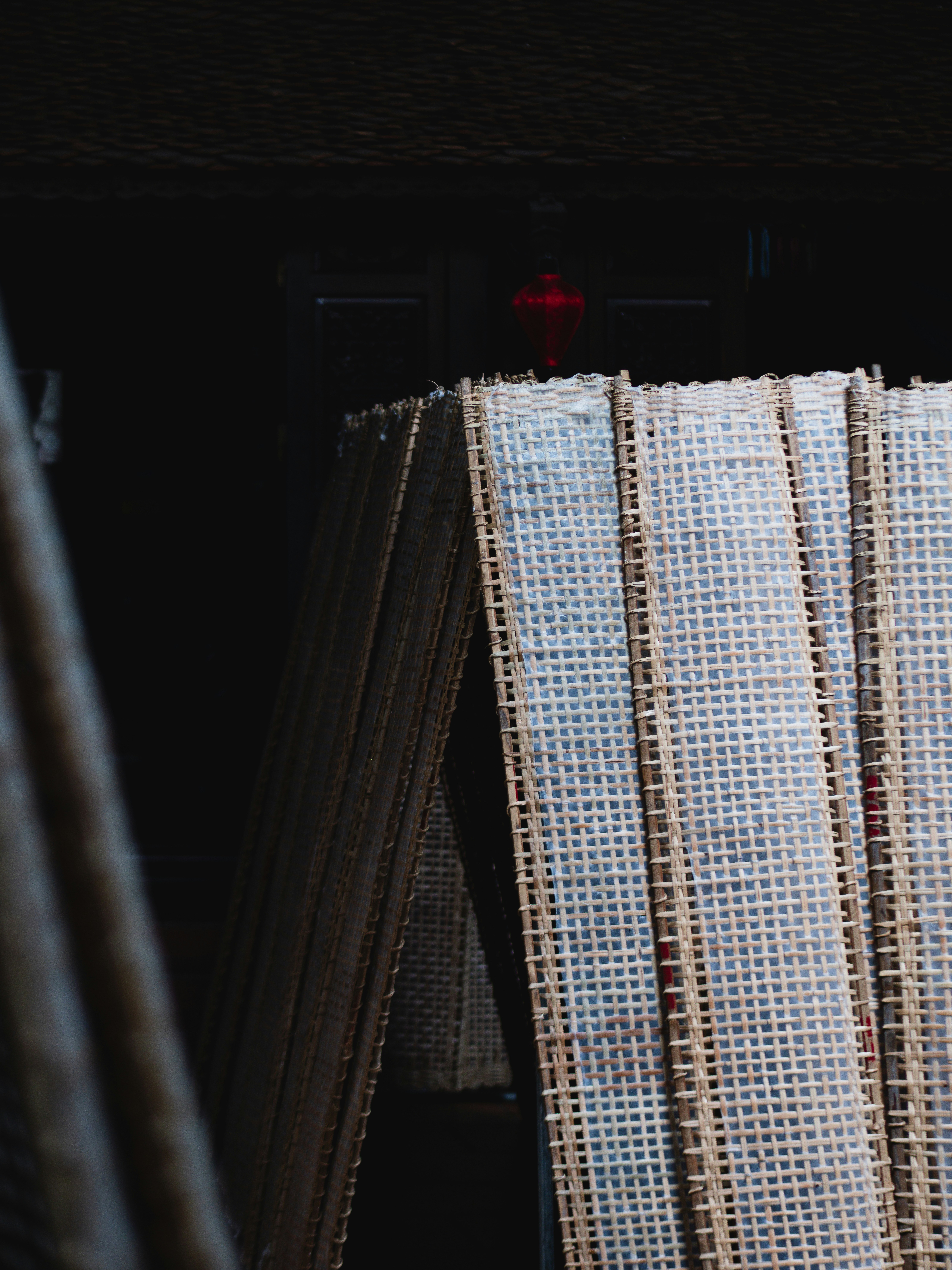 Close-up of woven cardboard or reed panels leaning in a dark workshop, forming vertical repeating textures with soft light highlighting the edges. Focus on material, depth, and industrial setting.