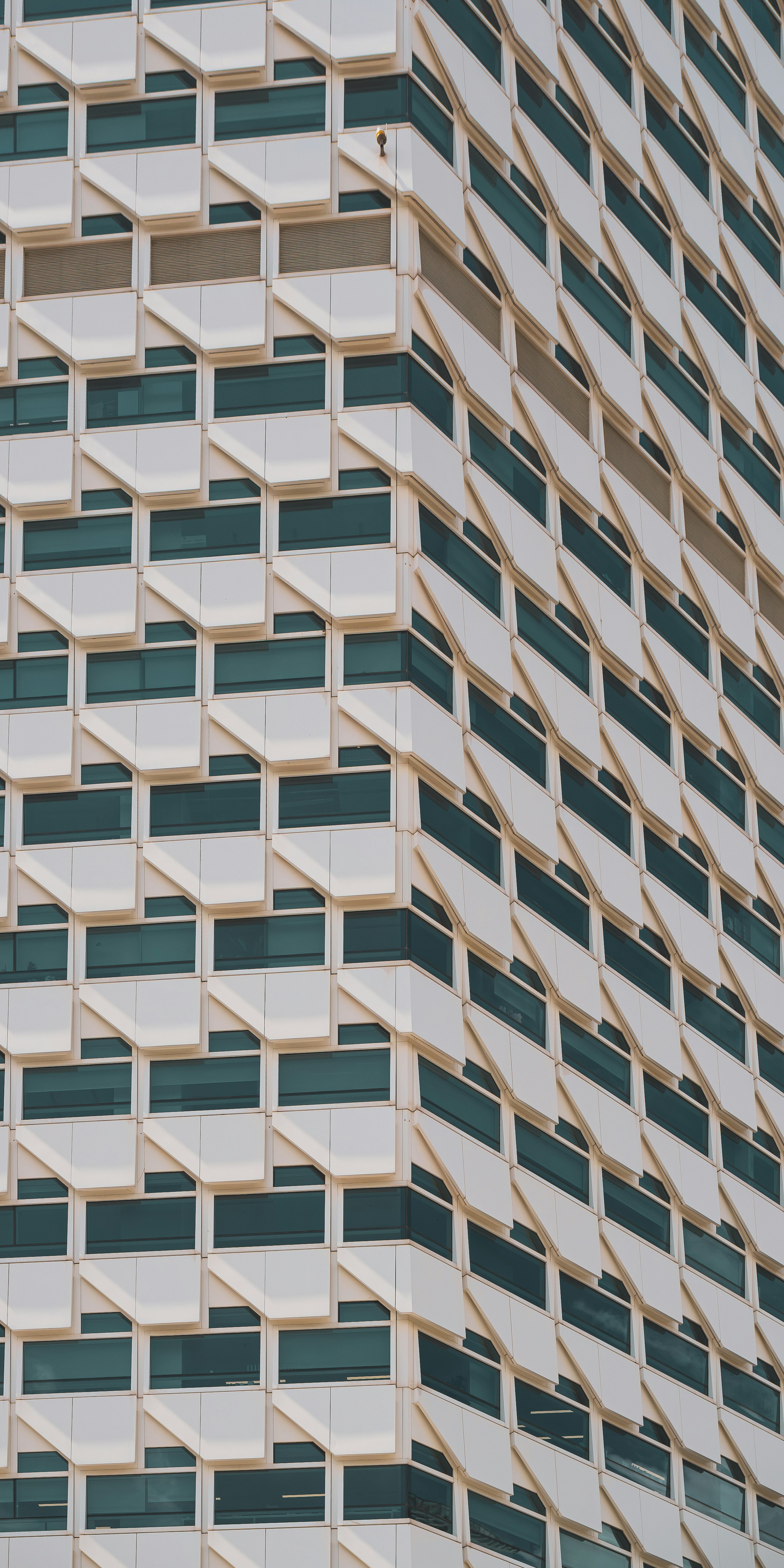 Geometric office building facade with a diagonal rhythm of repeating window bays and bevelled frames, highlighting light shadows and teal glass panes.