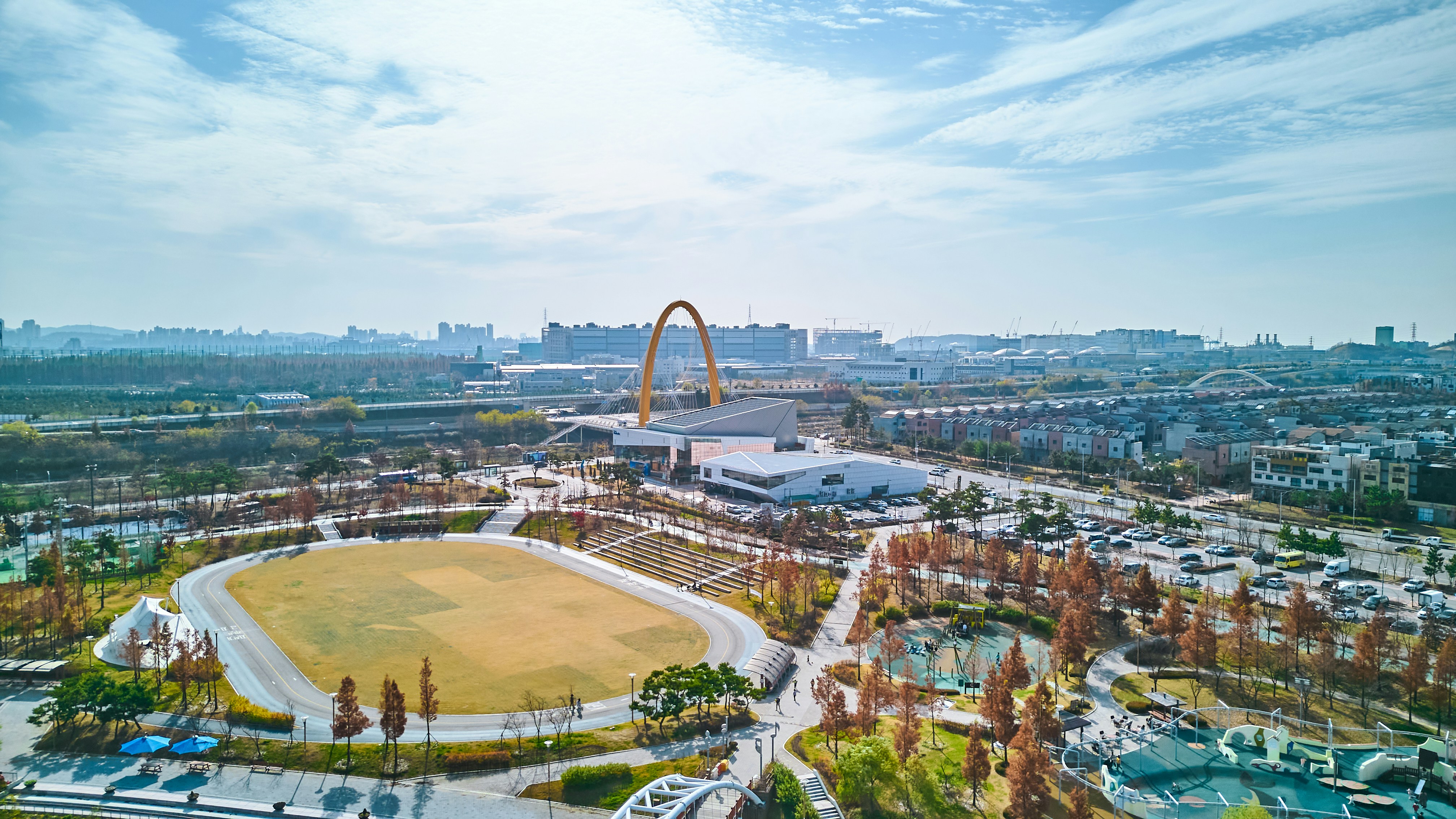 Aerial view of a modern urban park with a central athletic field, winding paths, and a distinctive arch structures amid trees and buildings.