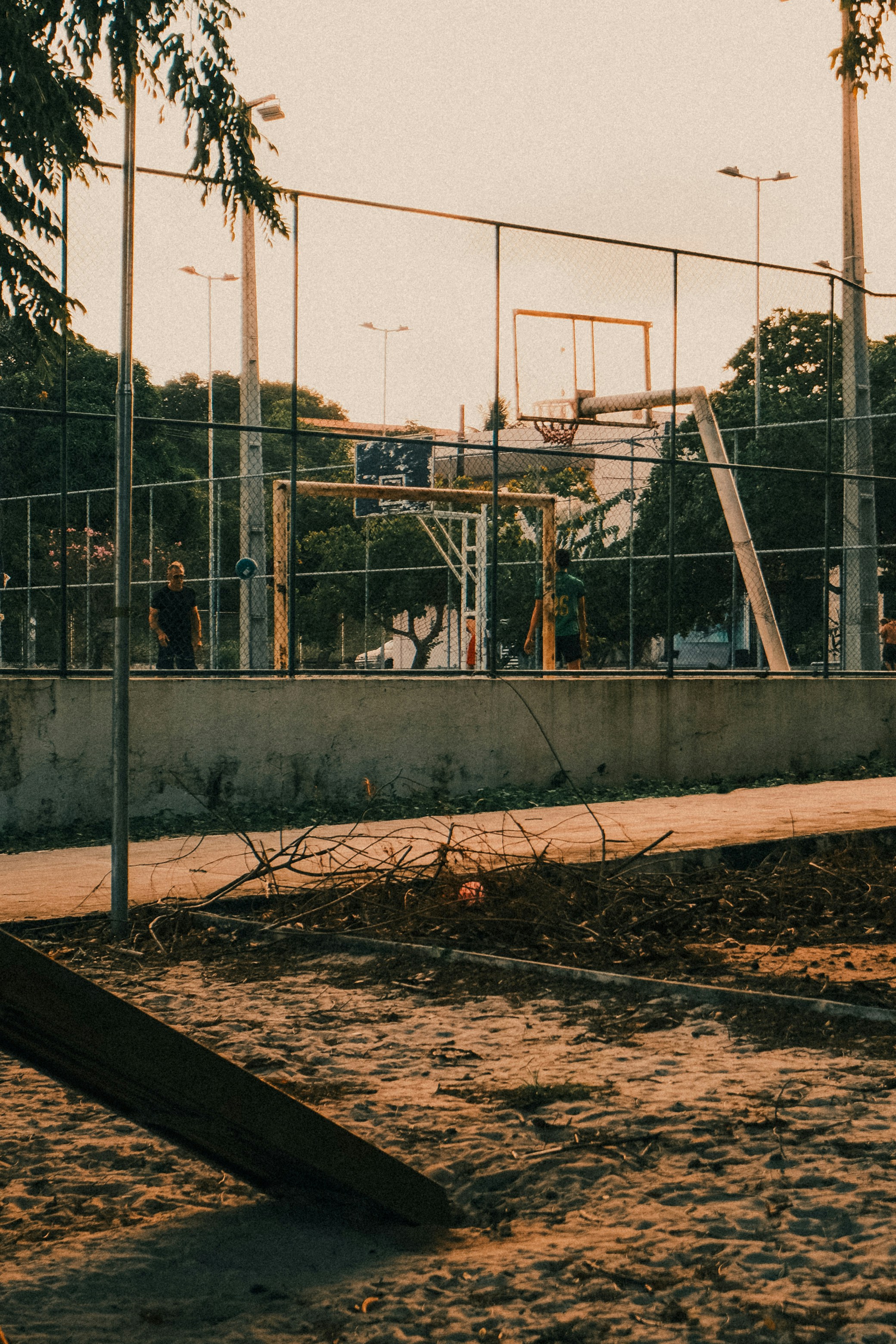 Basketball court behind a chain-link fence at dusk, with players and a hoop visible beyond the wall; warm, desaturated tones and a quiet, urban mood.