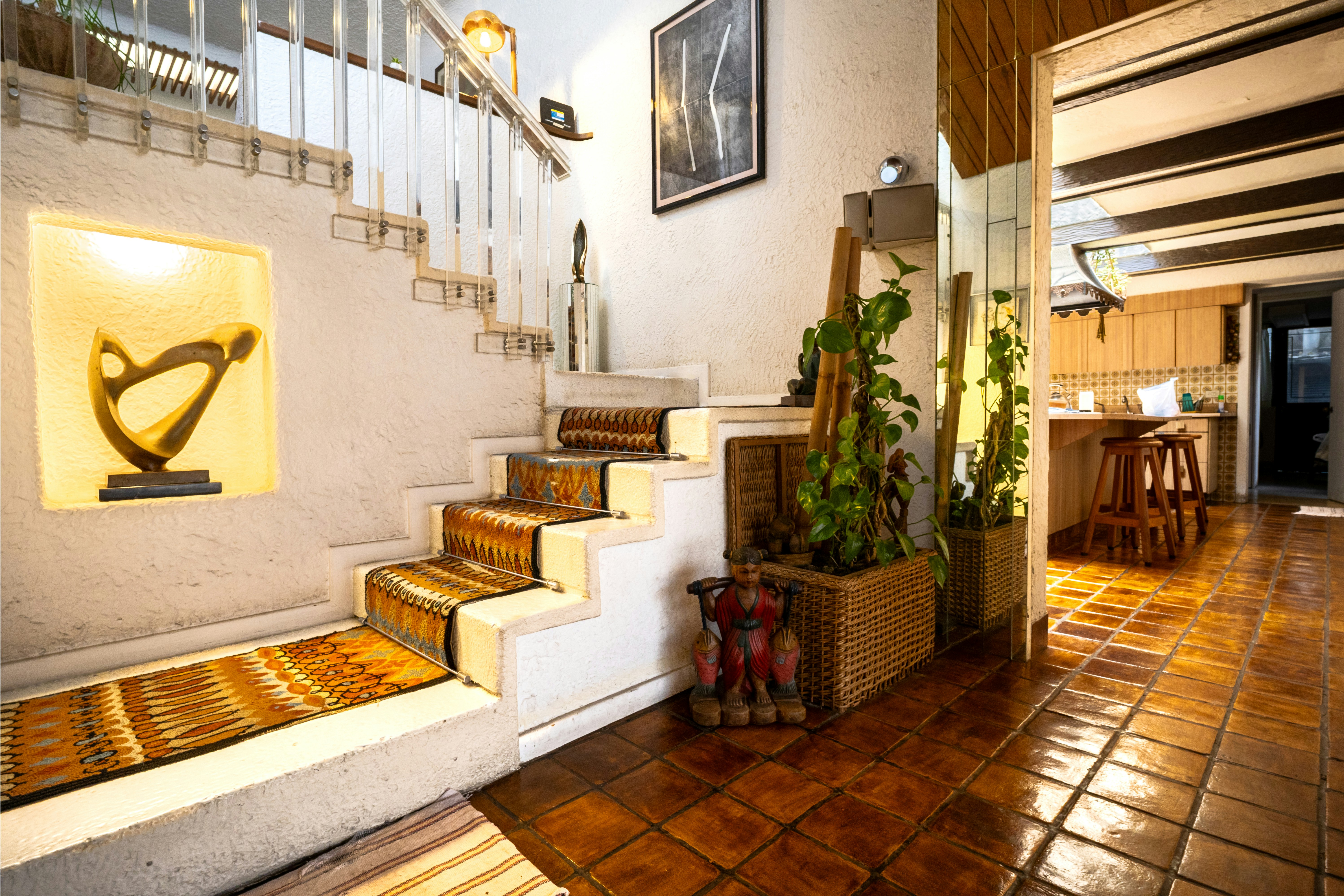 Warm-lit stairwell and tiled foyer with decorative art and greenery; a cozy, rustic interior space featuring a white staircase, woven runners, and tiled floor.