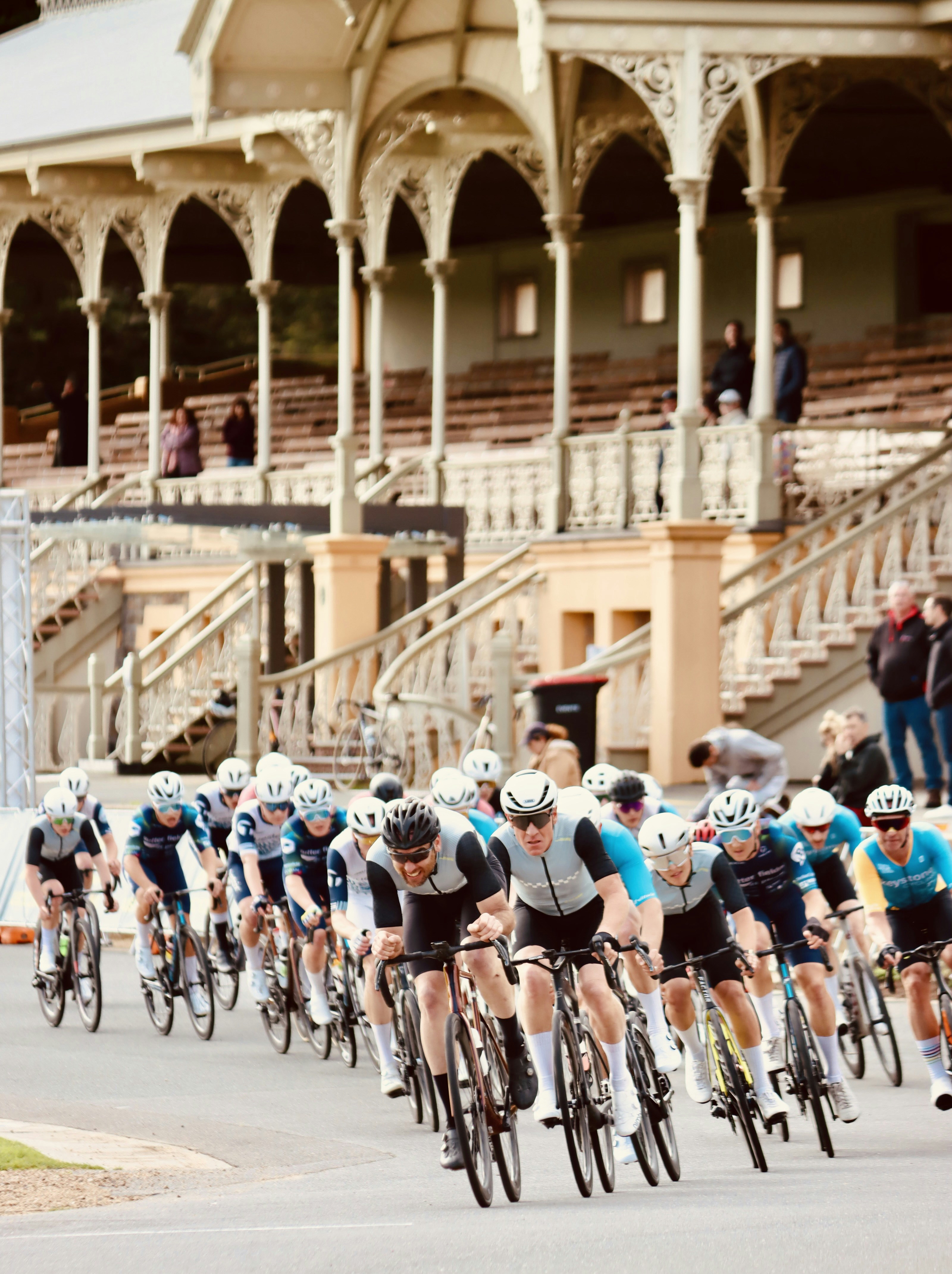 A pack of cyclists rocks through a curved street under ornate arches, riders in matching kits leaned forward in a race-like formation.