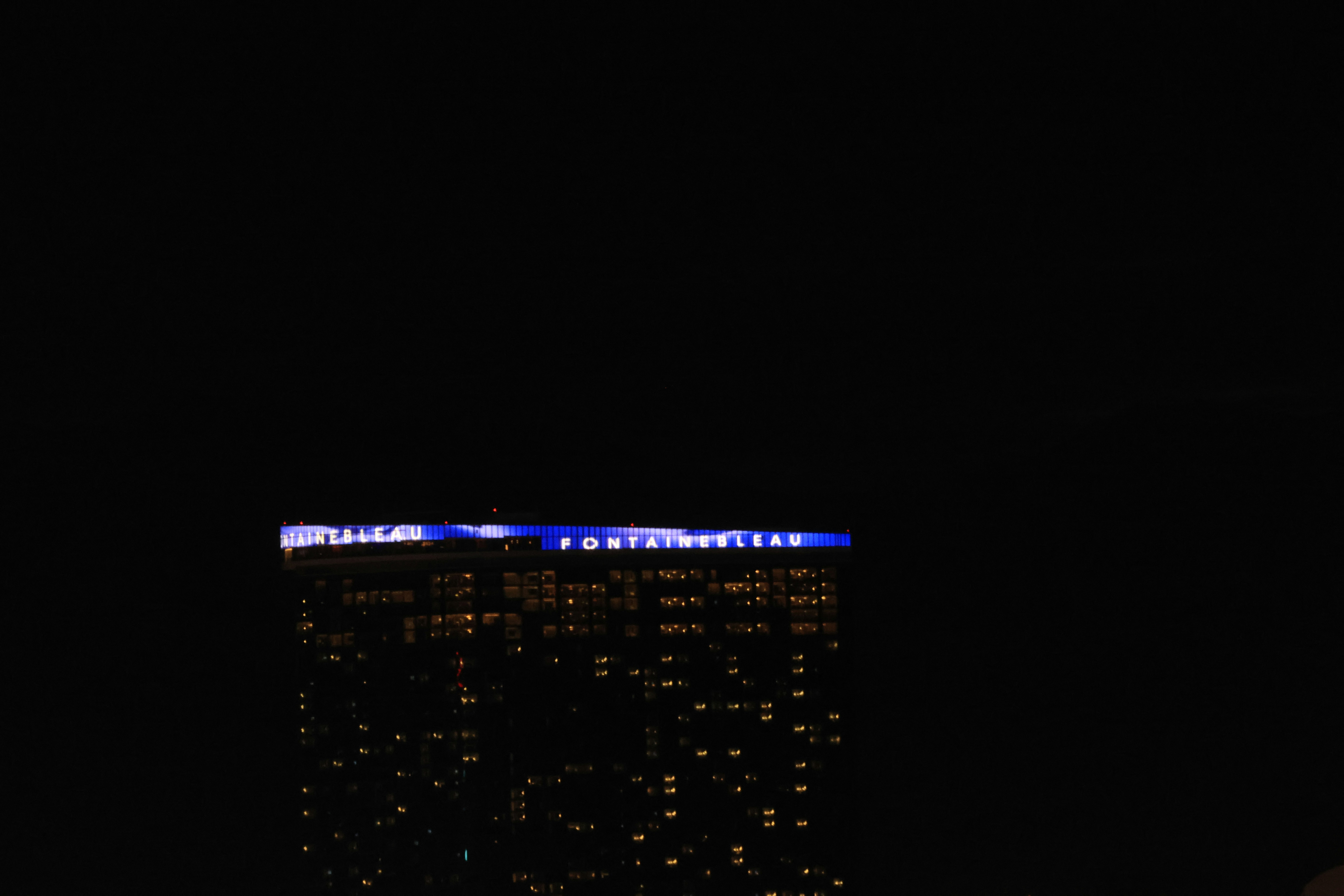 Tall building with illuminated sign at its top stands out against a dark sky, suggesting a nighttime cityscape. A logo or signage appears along the roof line, with lit windows creating a grid of warm points of light. (Logo visible at top edge.)