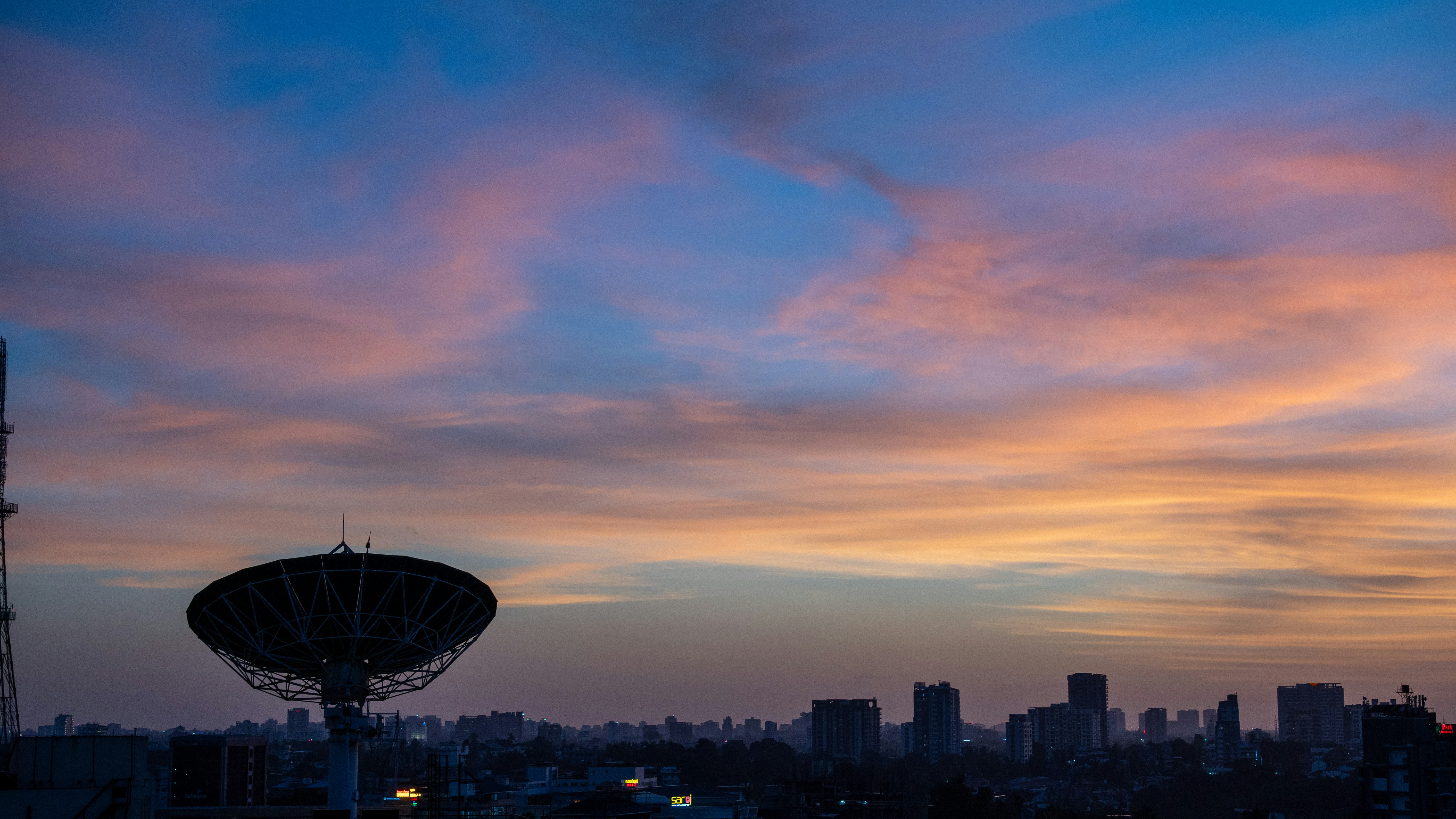 Silhouette of a large satellite dish against a city skyline at sunset, with warm pastel sky and distant buildings.