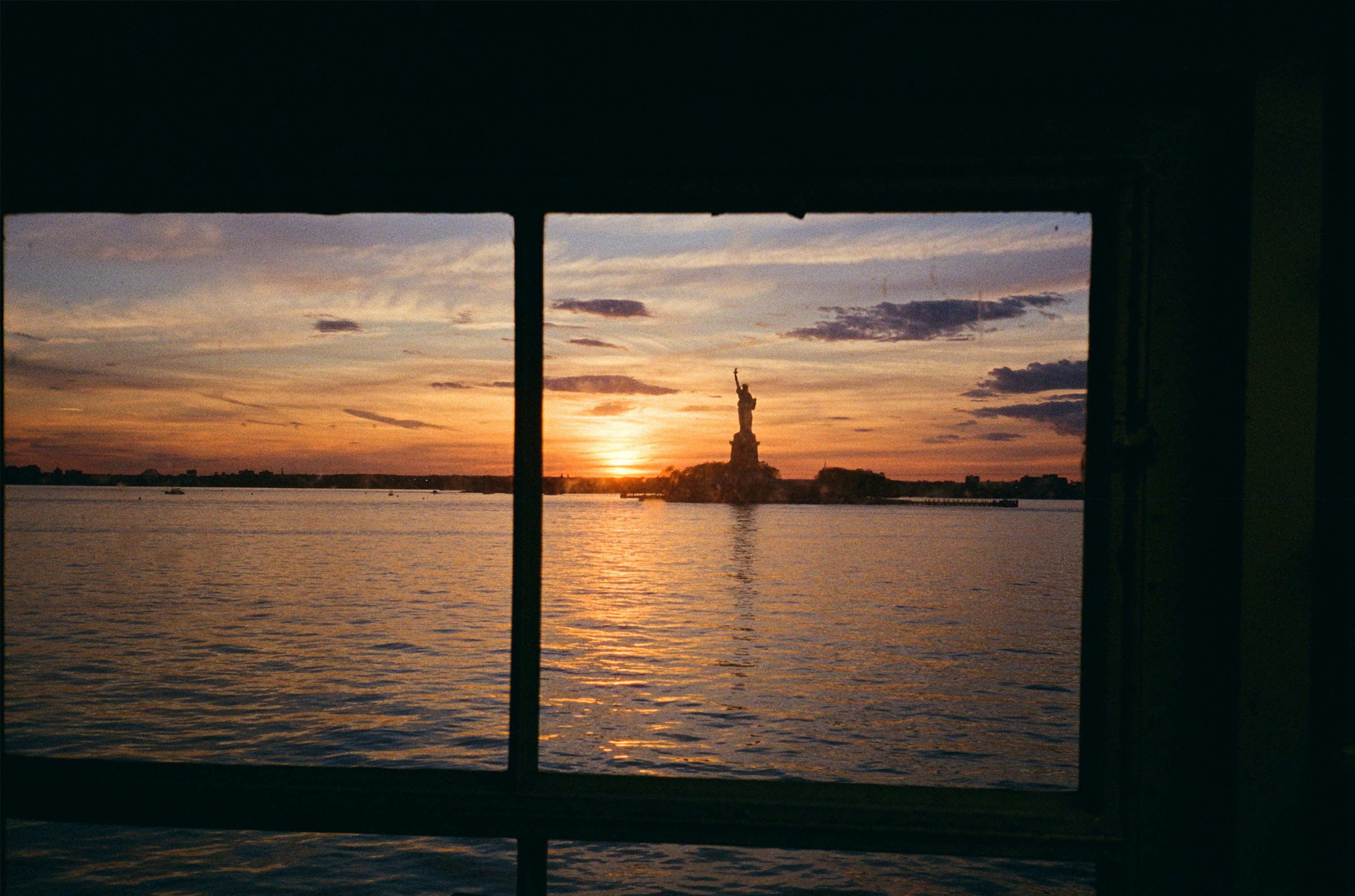Sunset over New York Harbor, seen through the window of a moving ferry. The Statue of Liberty is silhouetted against a glowing sky, while the last light of day stretches across the water in soft, shimmering reflections. Shot on an Olympus mju with Kodak Gold 200, capturing the warmth, grain, muted contrast, and nostalgic character of 35mm film.