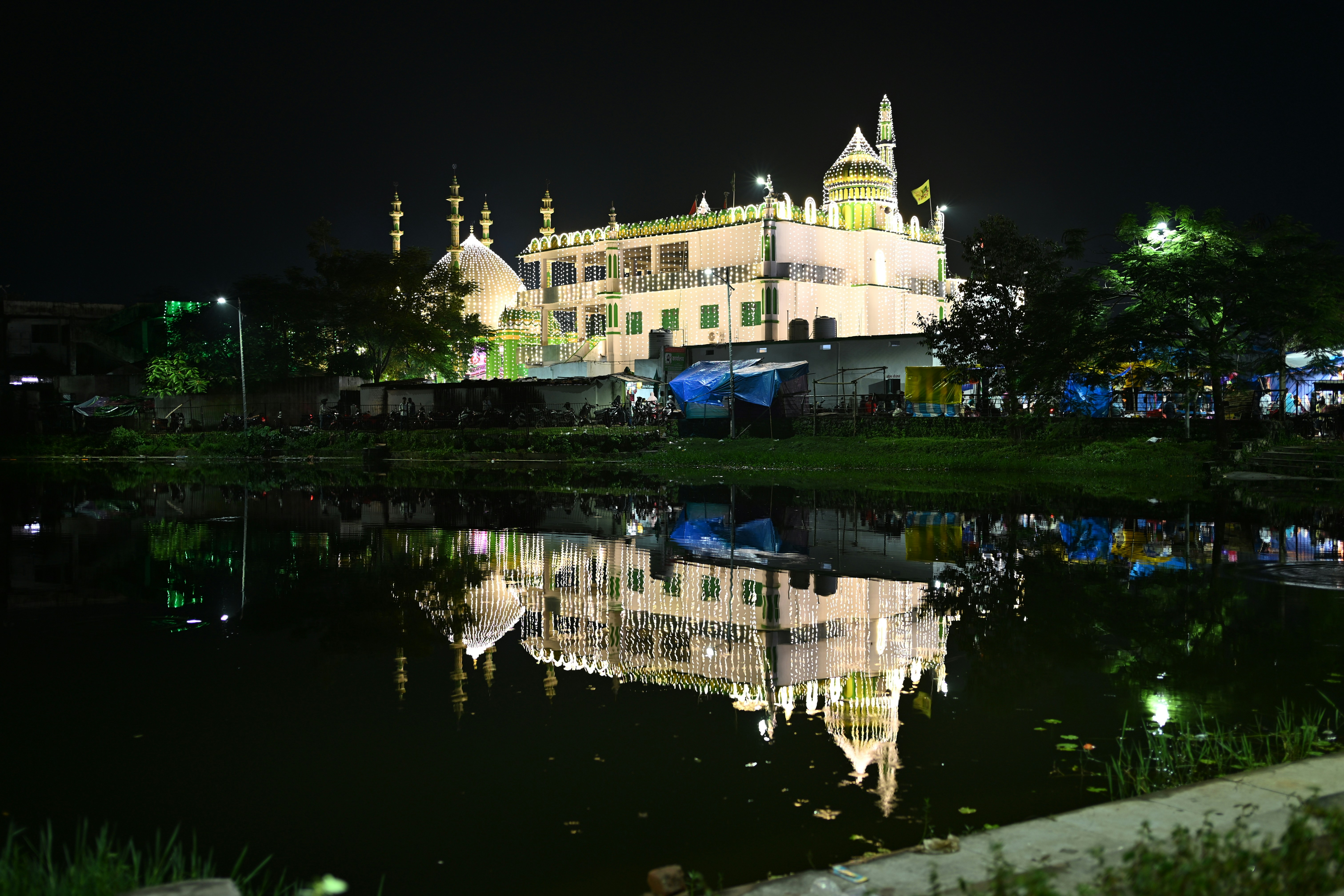 Night view of a lit temple complex along a calm river, with a clear reflection in the water and market tents at the base. A serene, low-light cityscape with soft greenery framing the scene.