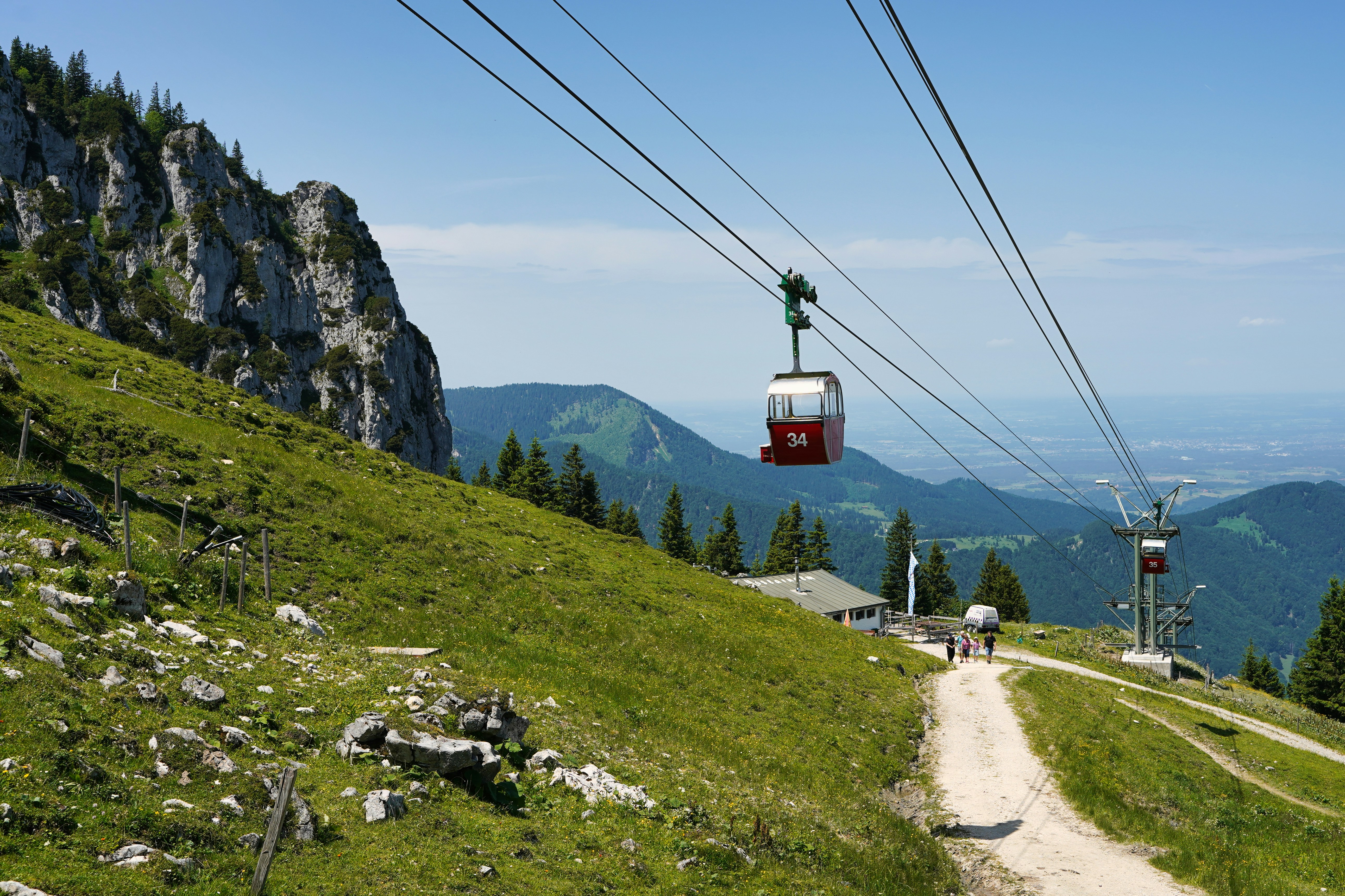 A red cable car suspended above a green mountain slope with a dirt path