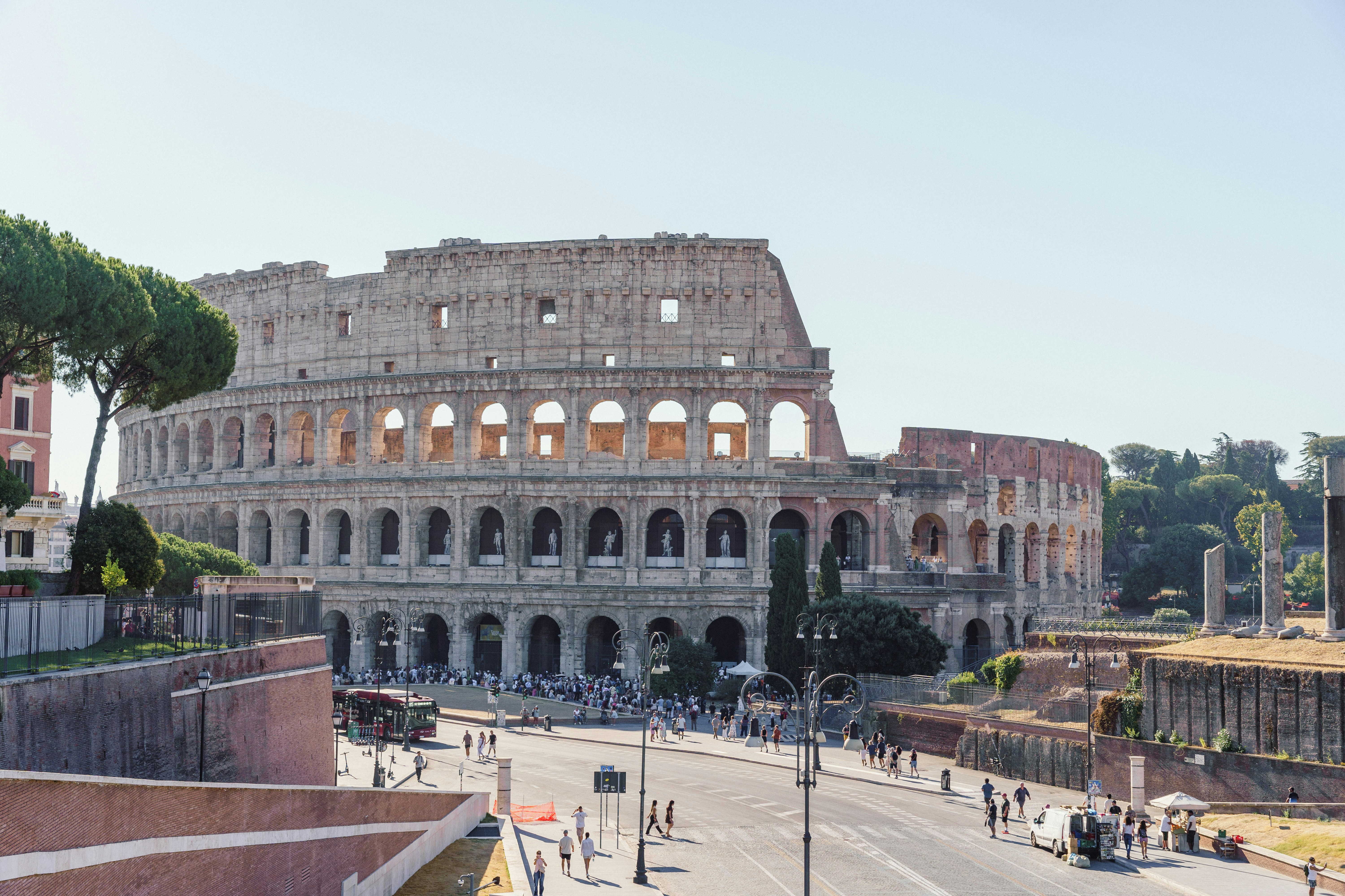 Historic Colosseum dominates the scene with visitors around the lower level arches and a sunlit plaza in front. Note: photograph captures a classic travel moment of the ancient amphitheater setting with onlookers and surrounding infrastructure.