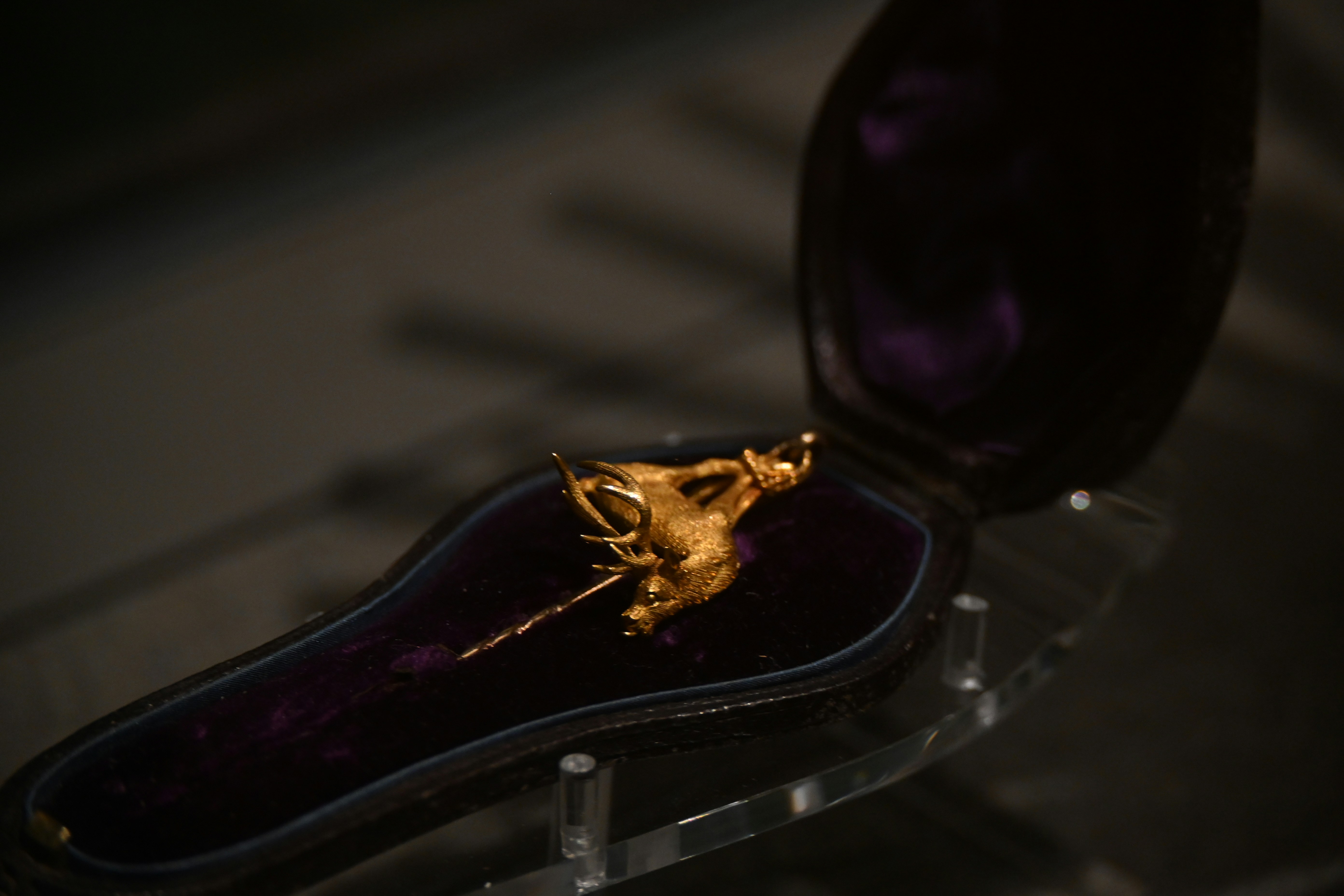 Close-up of a gold ring resting in a dark velvet-lined case, with subtle lighting highlighting metallic texture. The image focuses on a single object with moody, low-key tones and a hint of purple reflection in the case.