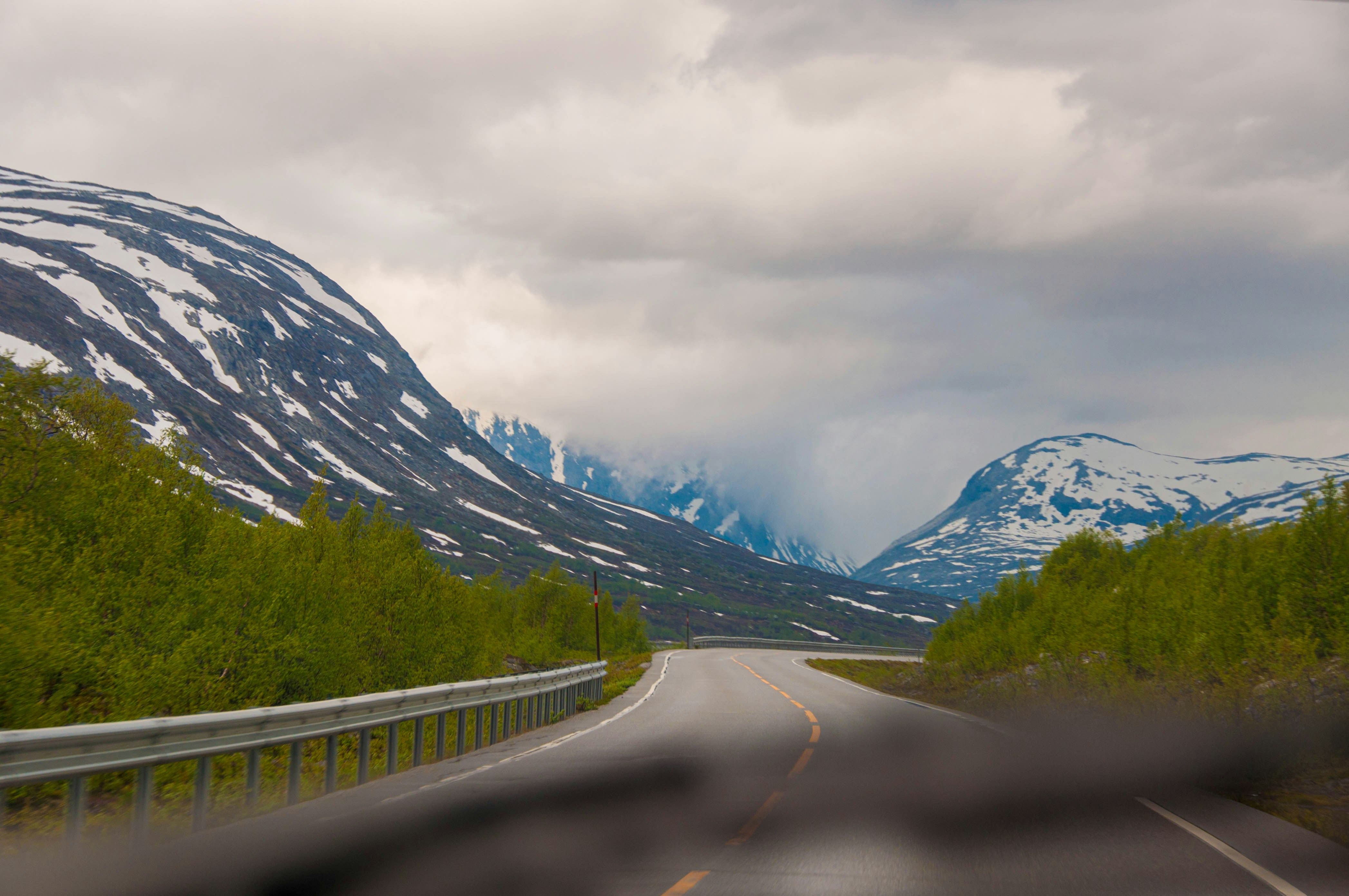 Snow-dusted mountains frame a winding road through a green valley, with a guardrail guiding the way and a blurred foreground suggesting motion.