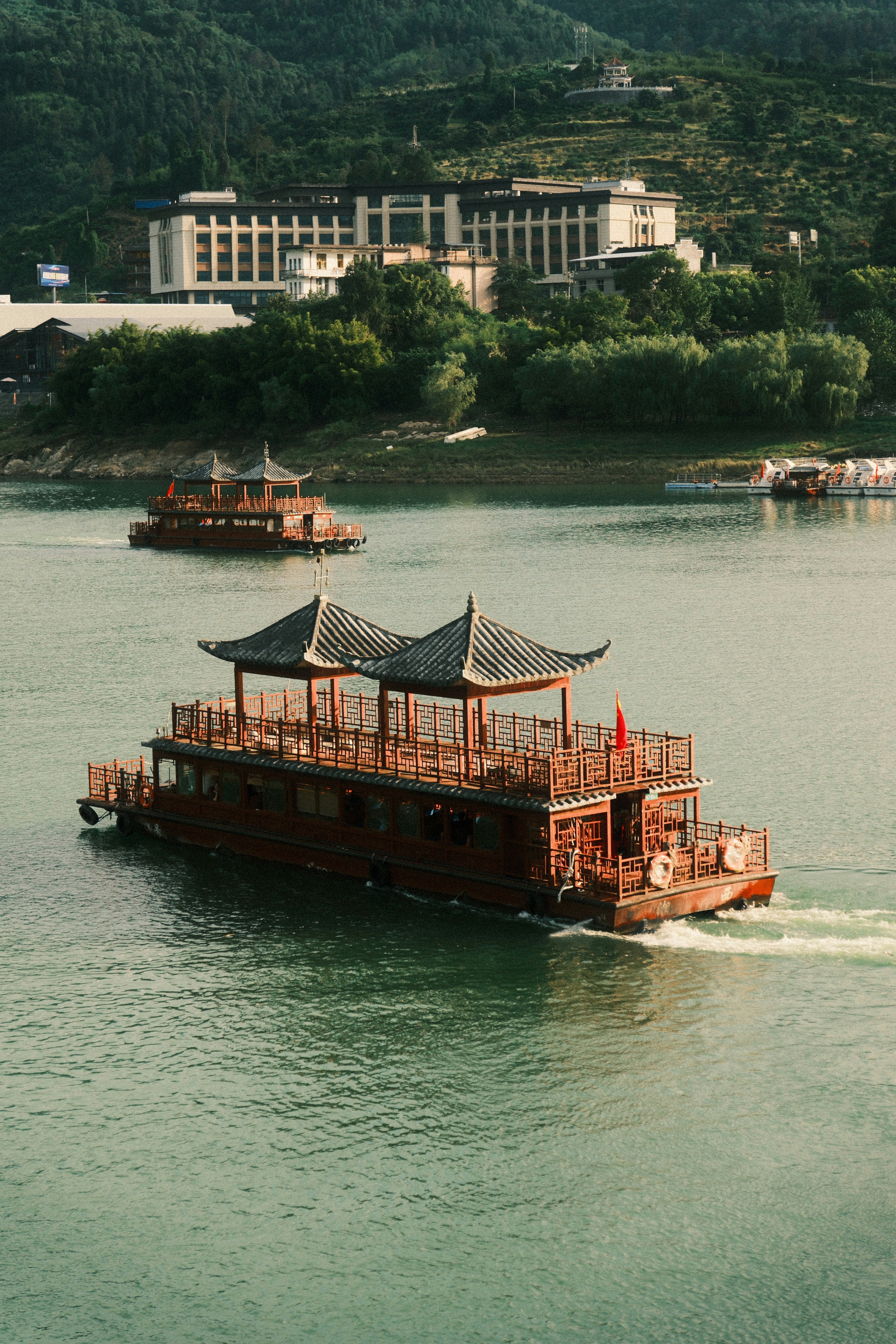 Red-hued riverboat gliding past a tree-lined bank under a hillside complex