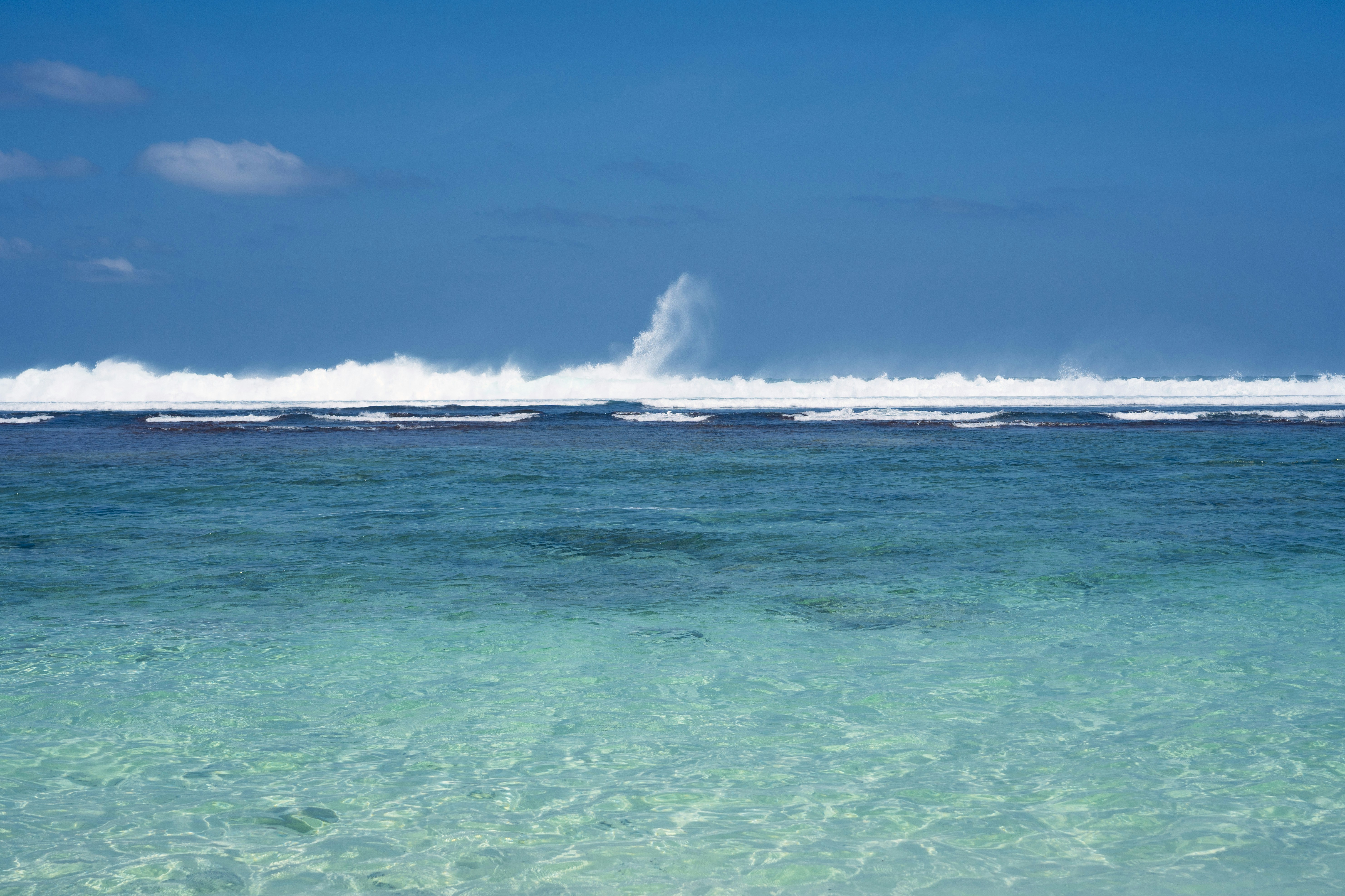 Bright turquoise shallows meet a deep blue horizon as rhythmic waves crest on a sunlit ocean. A tranquil seascape emphasizing clear water, gentle gradients, and a minimal sky.